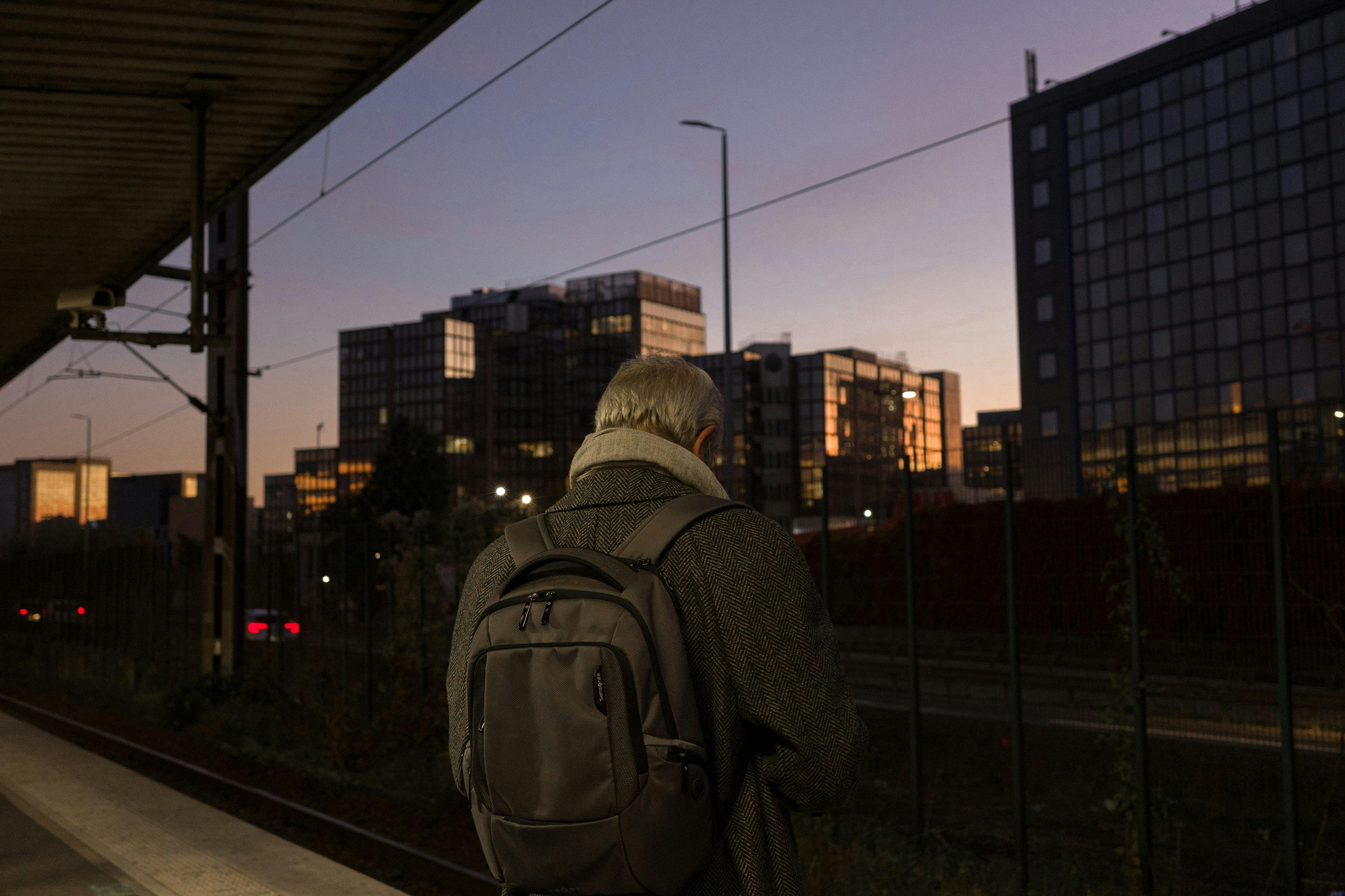 Person with a backpack stands on a train platform at dusk, with city buildings illuminated in the background.