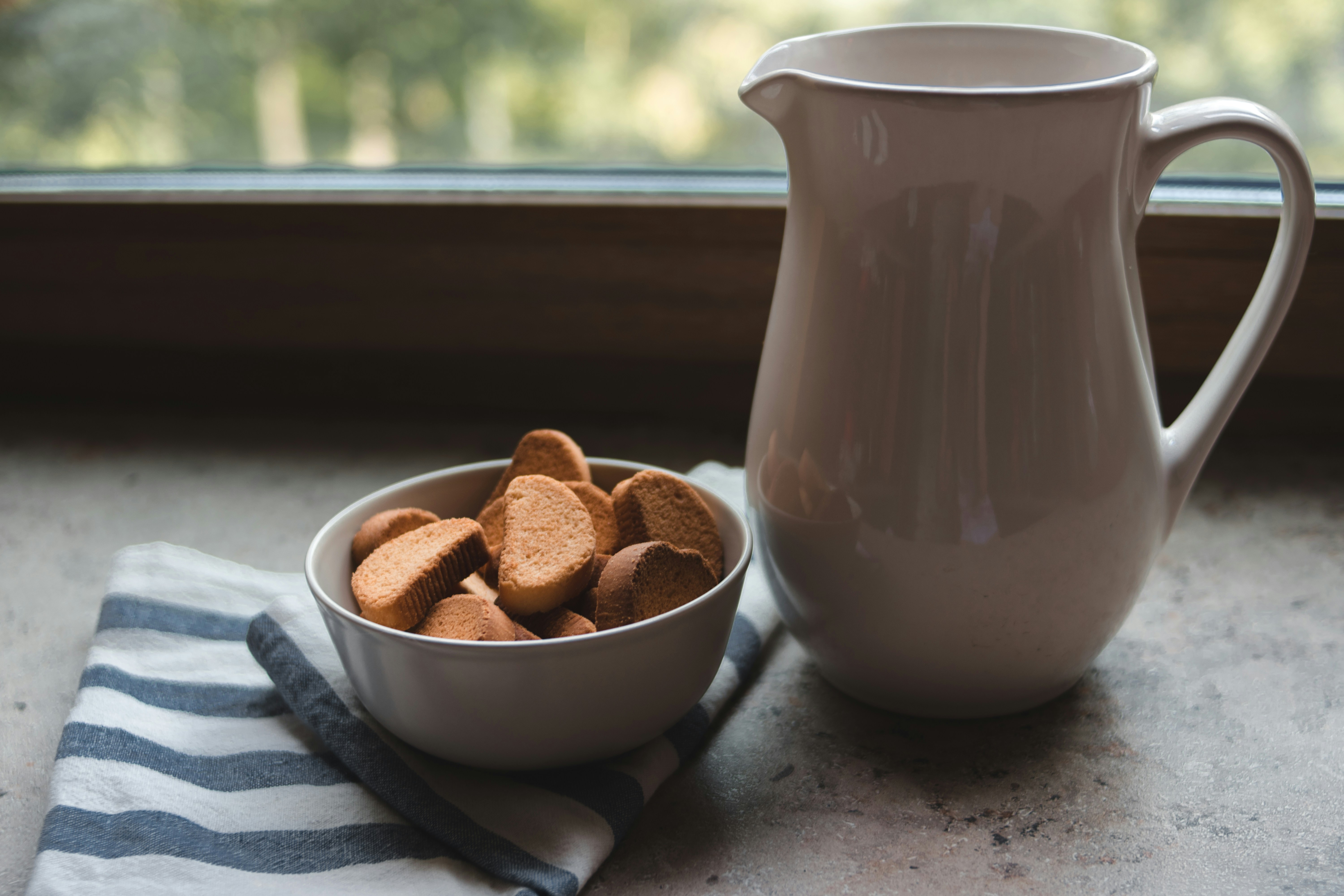 A white pitcher sits beside a bowl of assorted cookies on a striped cloth, illuminated by soft natural light from a nearby window.