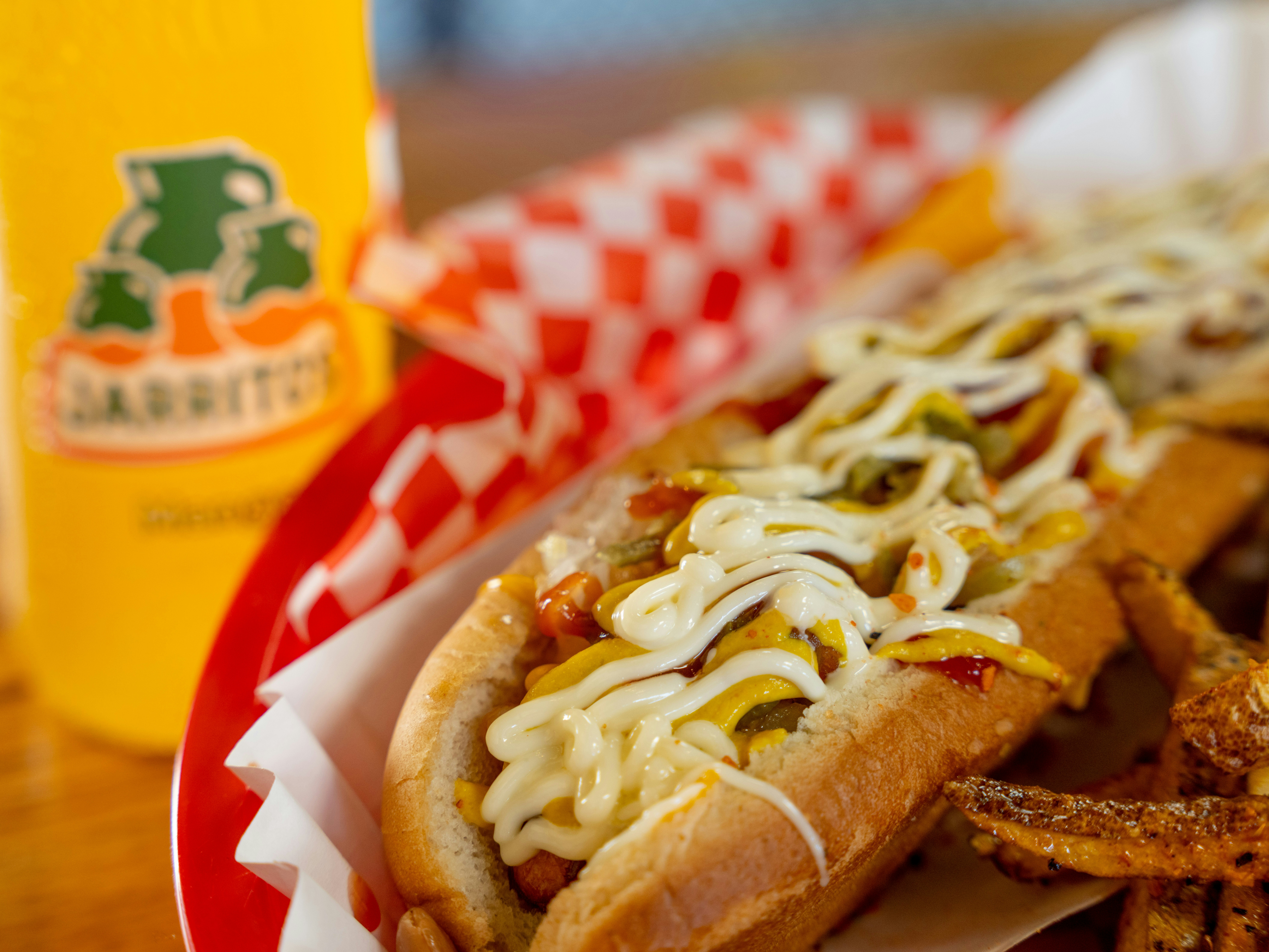 Delicious hot dog topped with mustard, ketchup, and mayonnaise, accompanied by a side of crispy fries and a refreshing drink in the background.