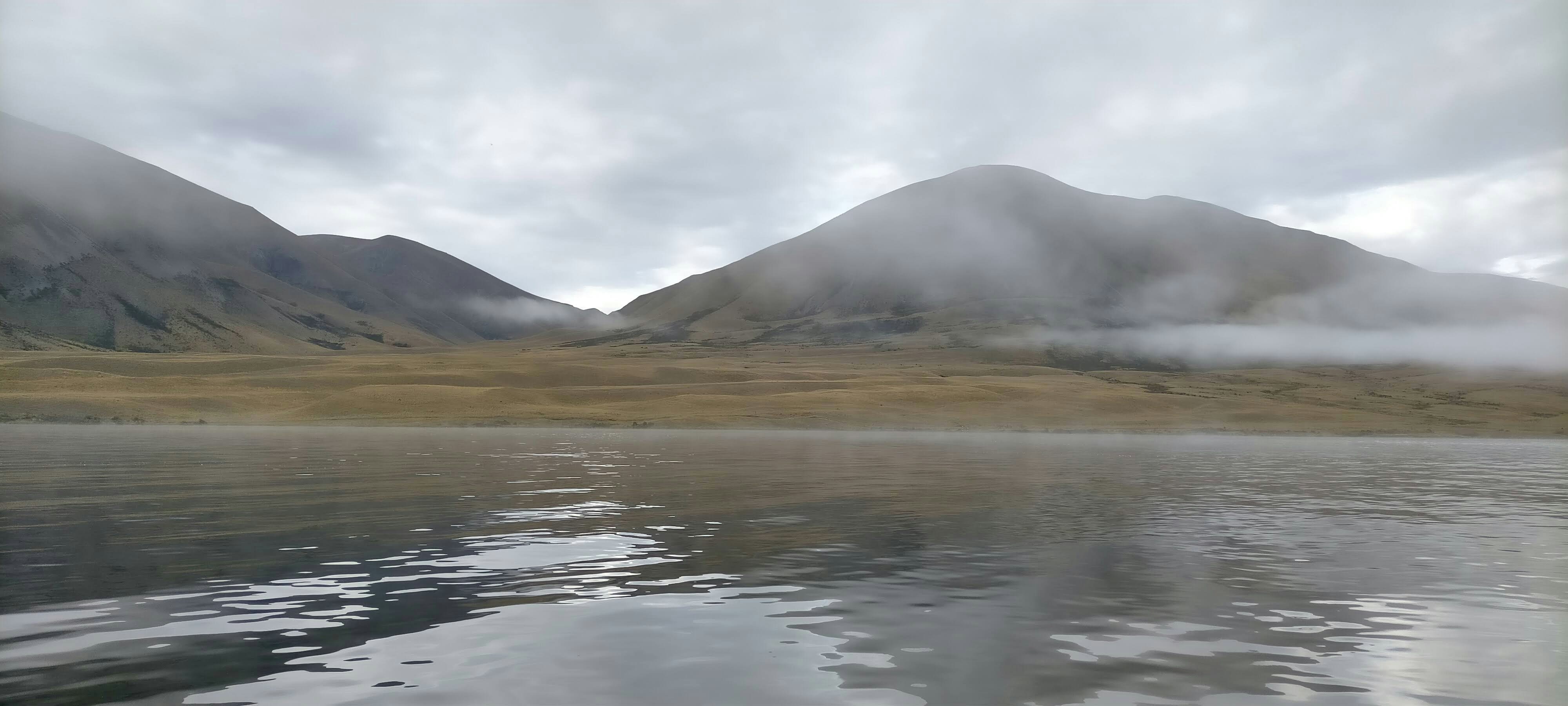 Calm lake reflecting misty mountains under a cloudy sky, creating an atmosphere of tranquility.