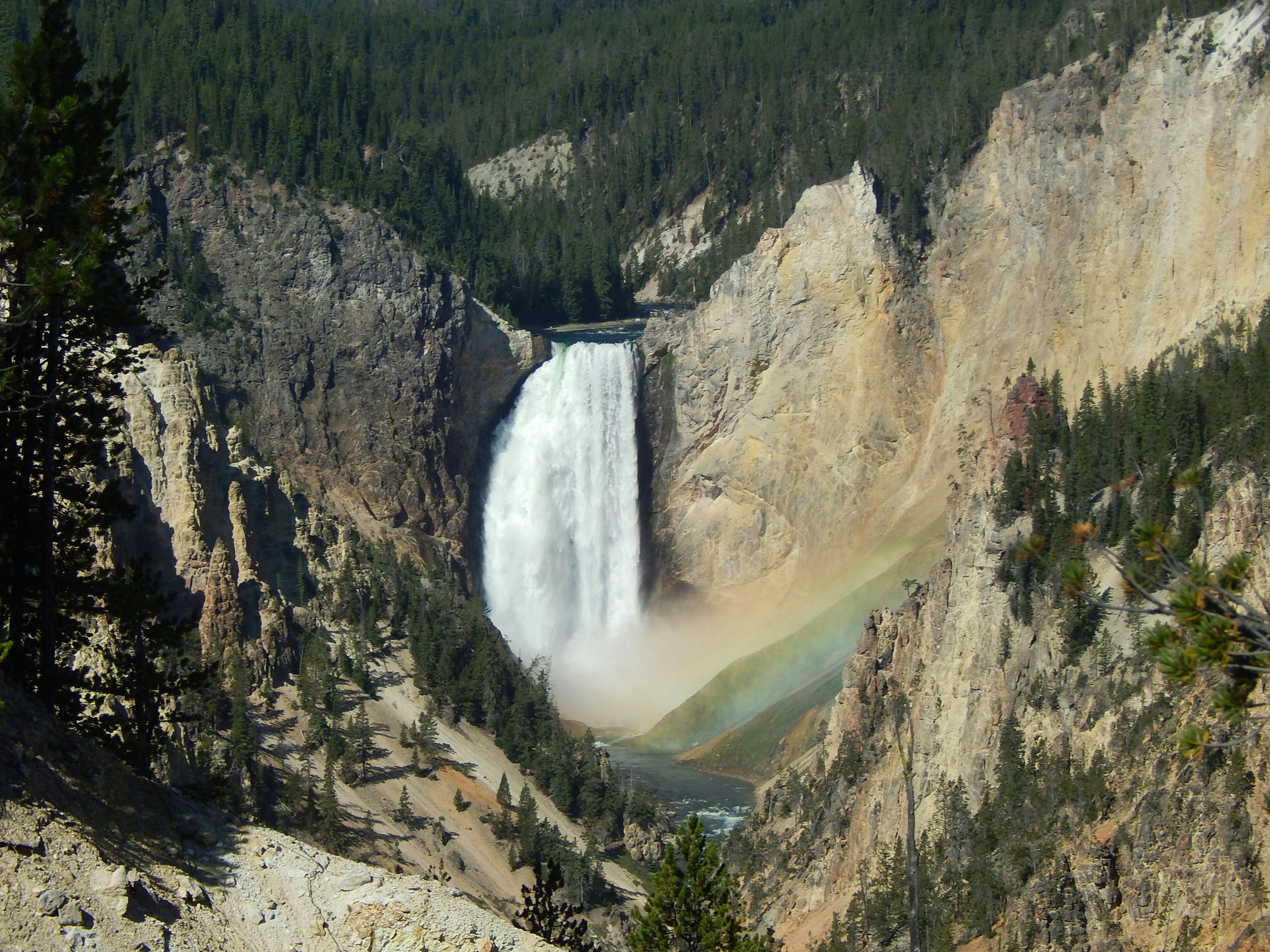Waterfall plunging into a canyon surrounded by rugged cliffs and dense forest with a faint rainbow at the base.