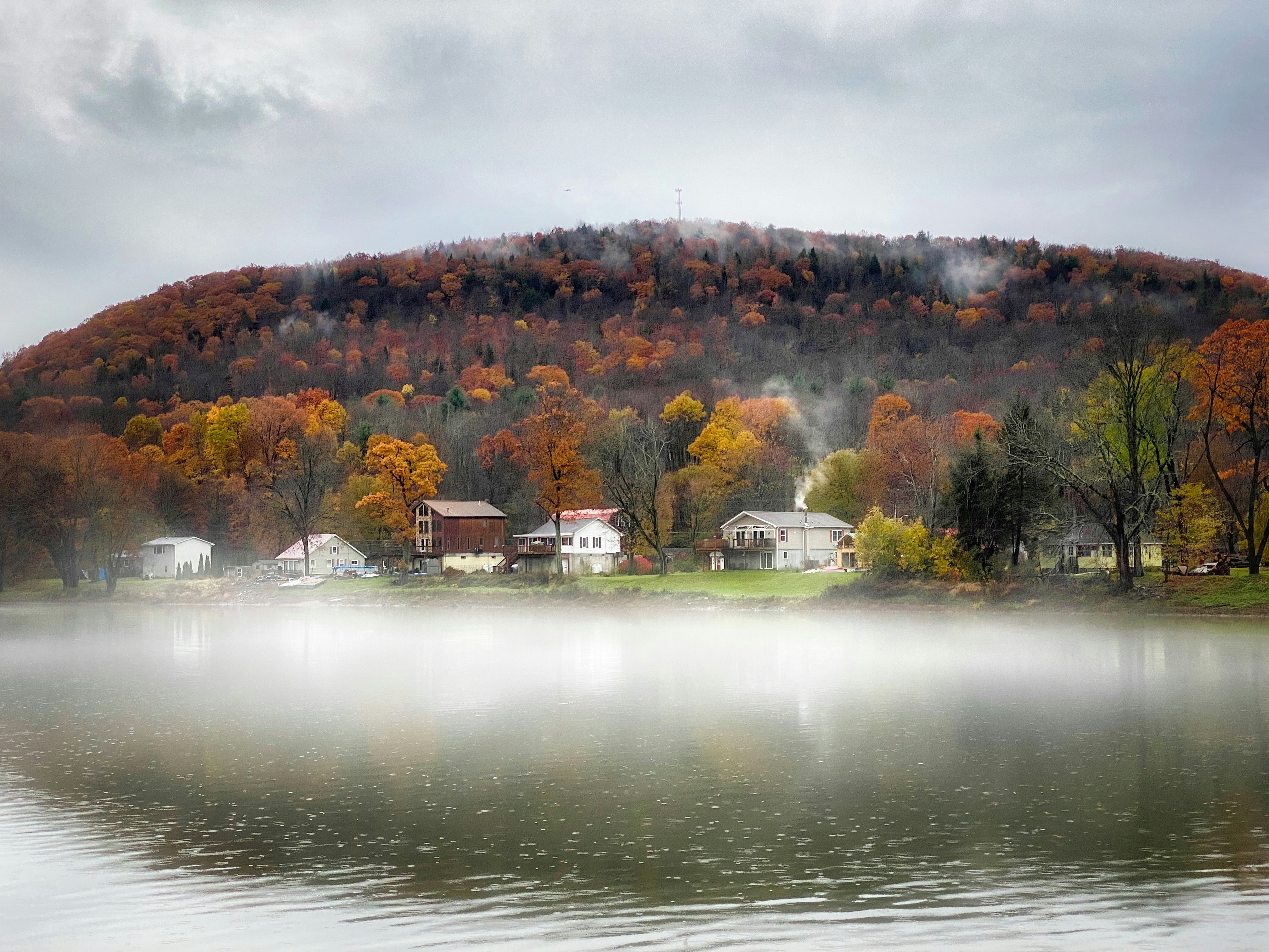 Fog drifts over a lake with vibrant autumn foliage and houses nestled beneath a wooded hill.