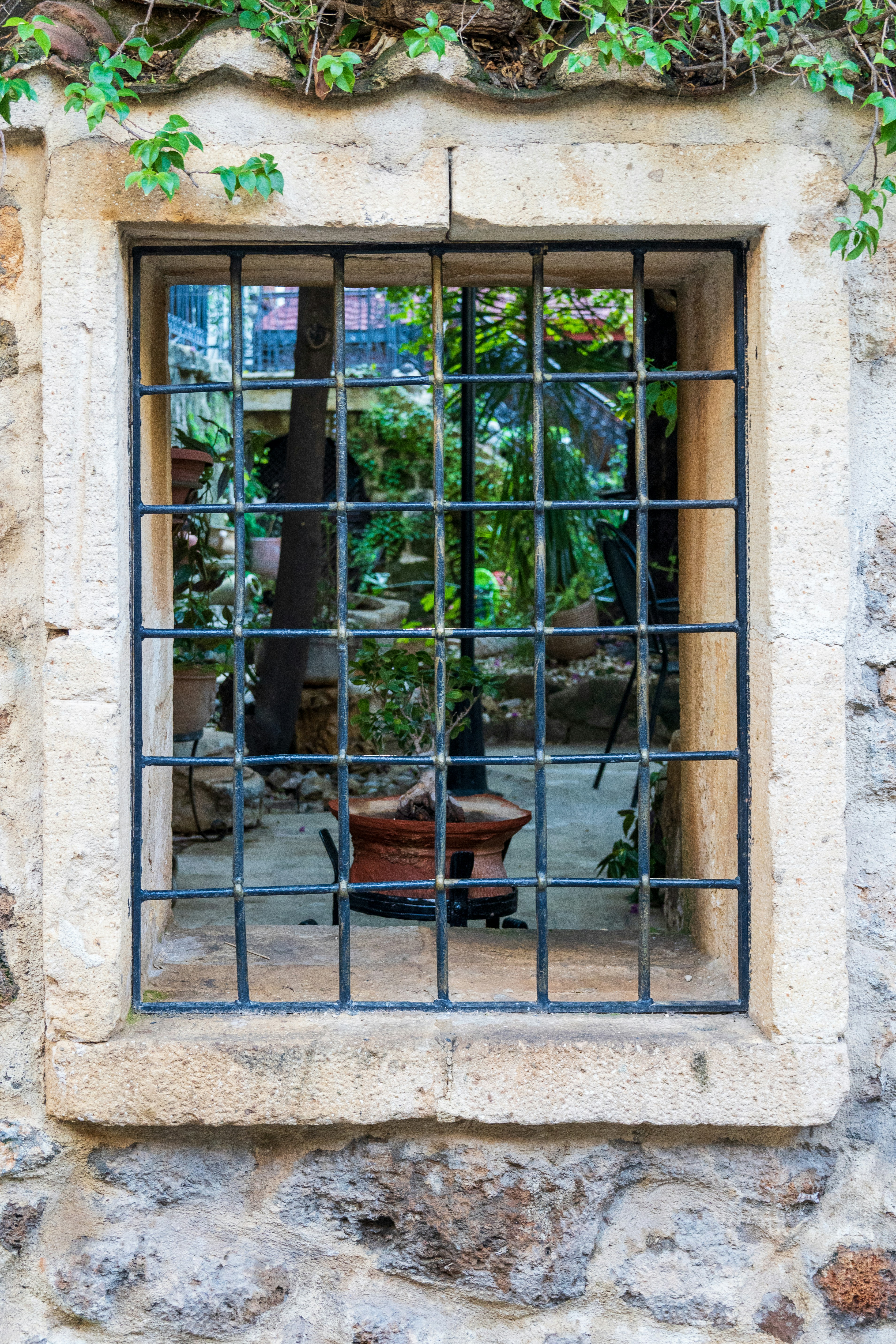 A rustic stone window framed by greenery reveals a serene garden scene with a potted plant at its center.