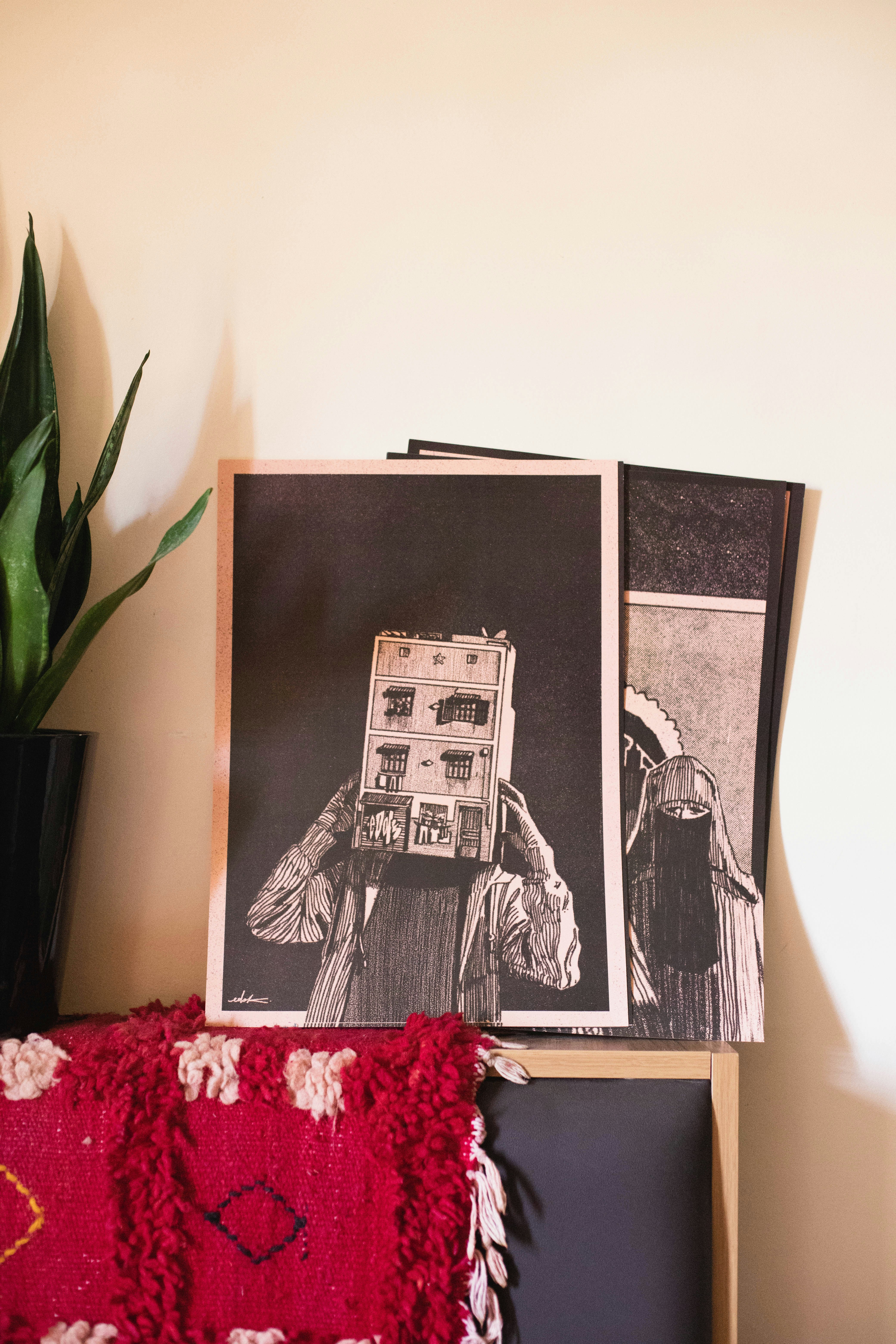 Artistic illustration depicting a figure holding a building facade, surrounded by layered artworks and a plant. A vibrant textile adds warmth to the scene.