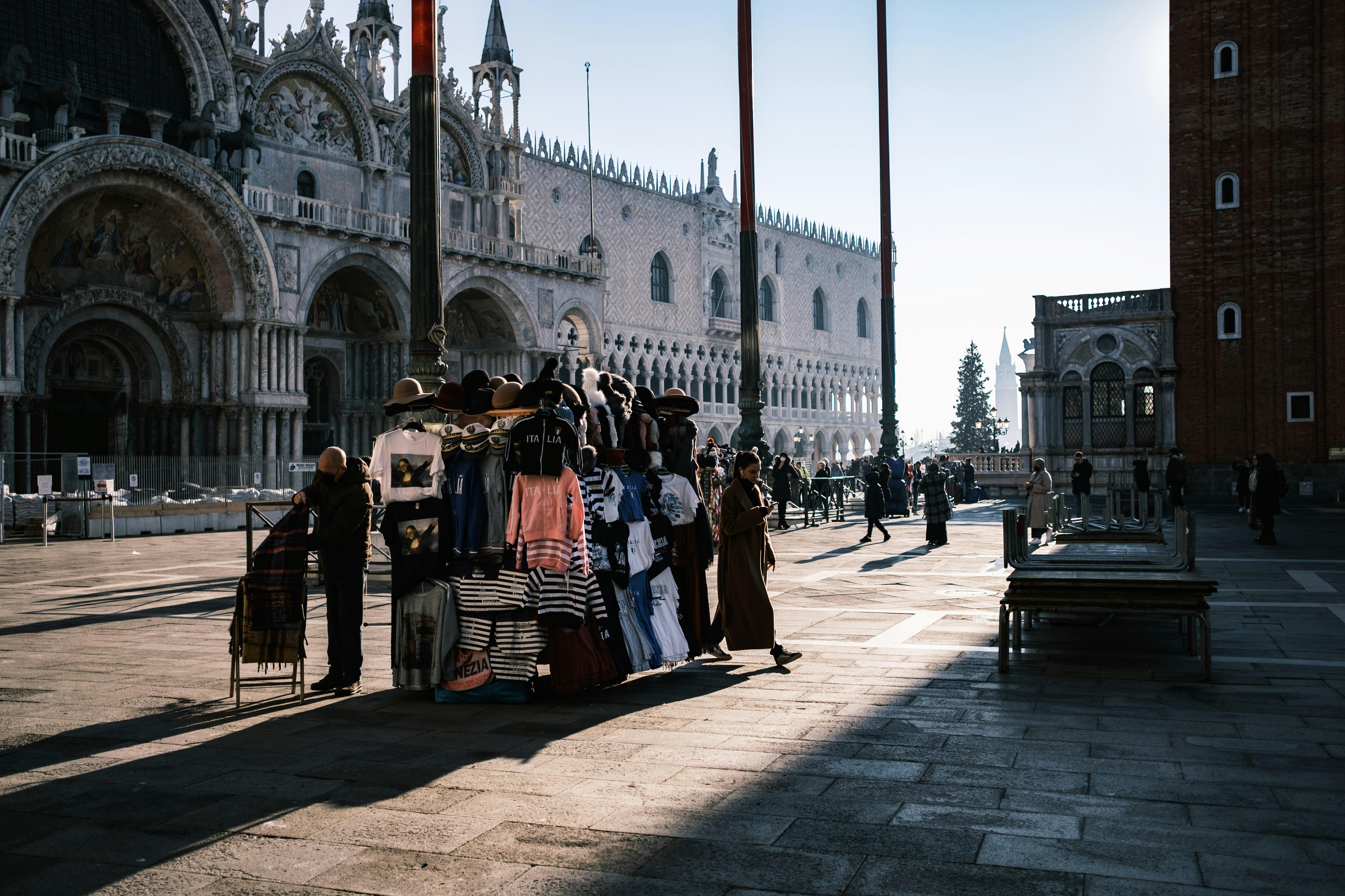 Street vendors with clothing racks casting long shadows in a sunlit Venetian square.