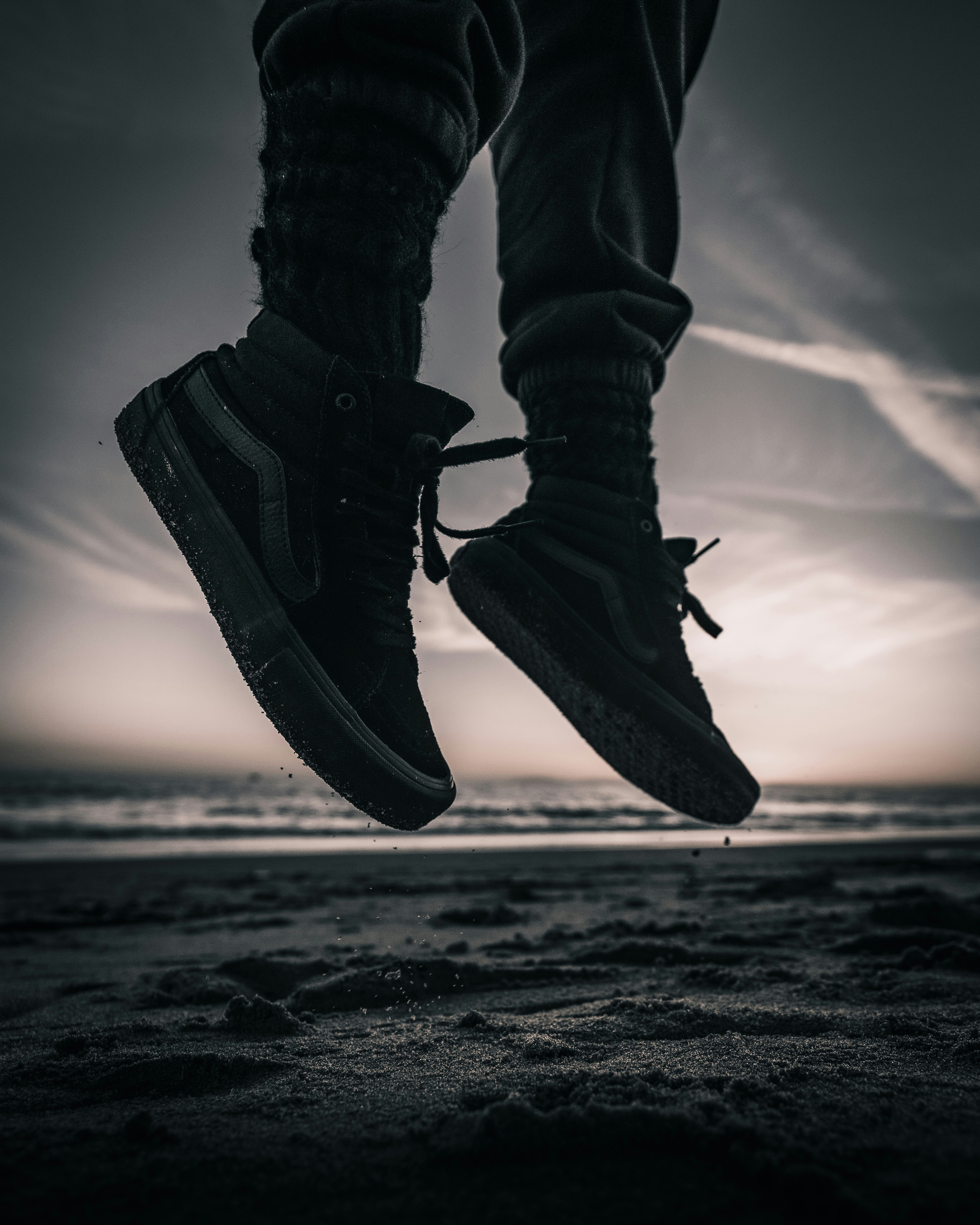 Black sneakers suspended mid-air against a moody beach backdrop at sunset, emphasizing motion and freedom.