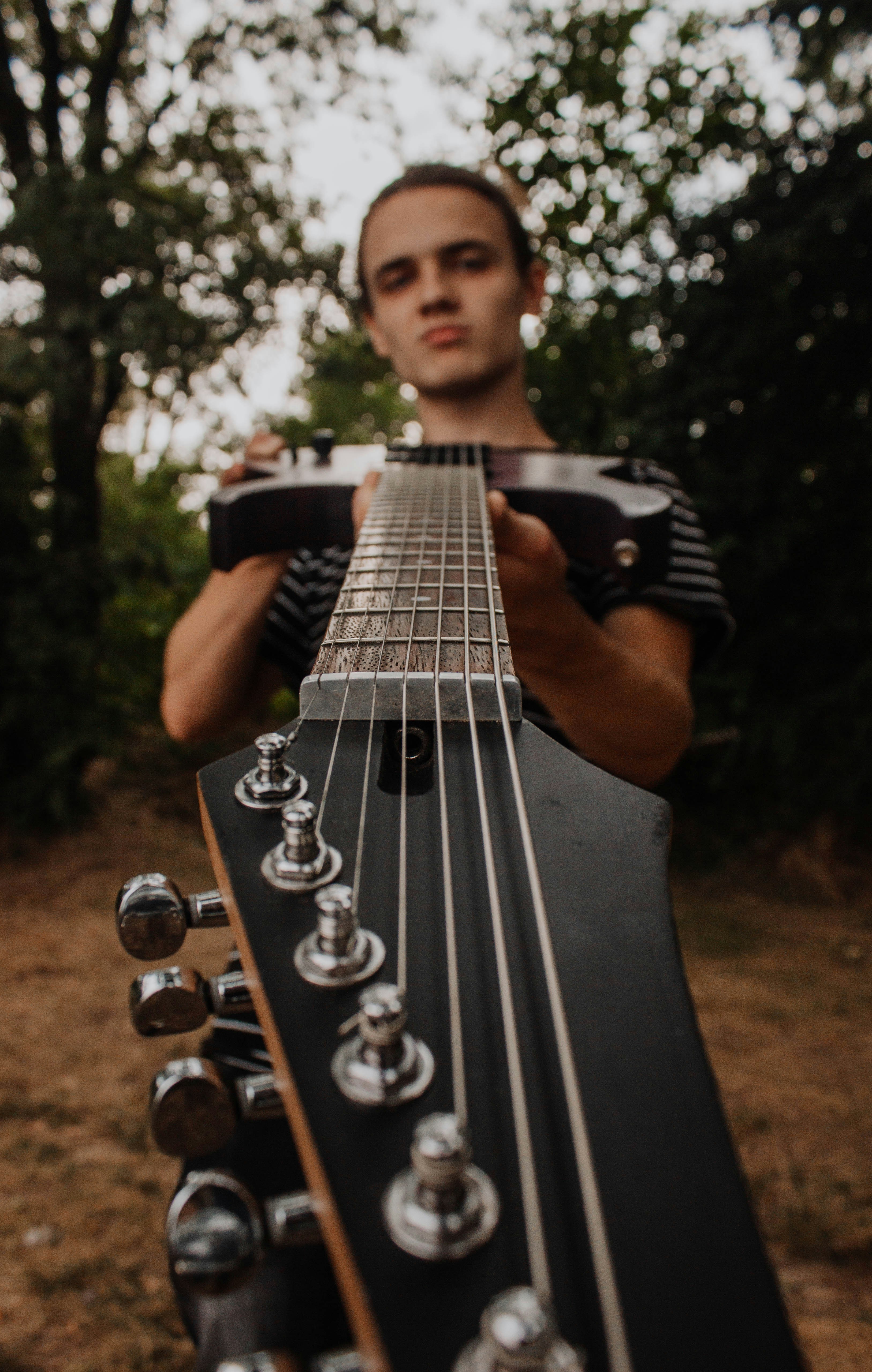 Close-up view of a guitarist's hands on the neck of an electric guitar, with a blurred background of greenery. The perspective emphasizes the instrument's strings and tuning pegs.