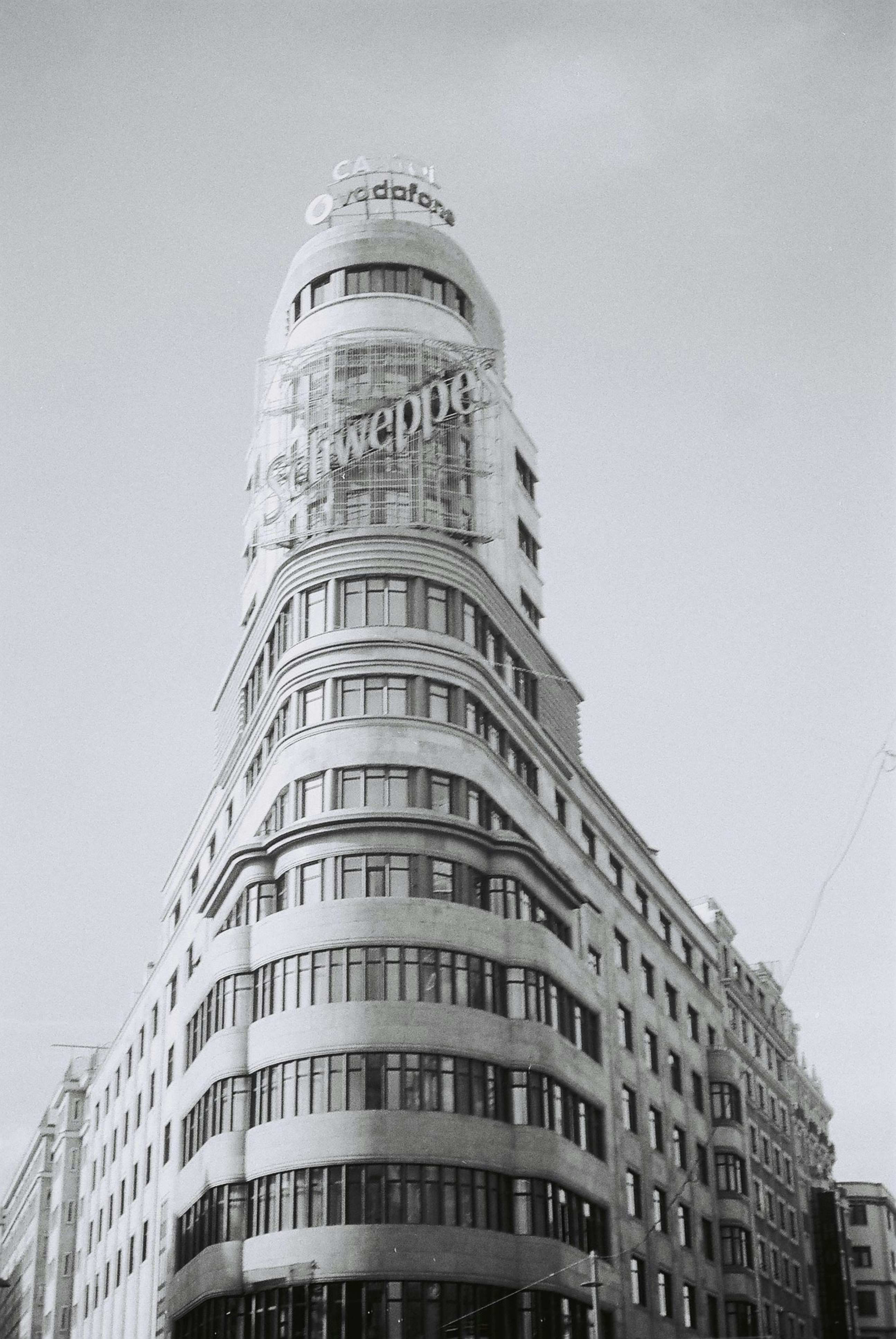 A monochrome photograph of a tall curved-corner office tower with a prominent façade sign and a grid of windows.