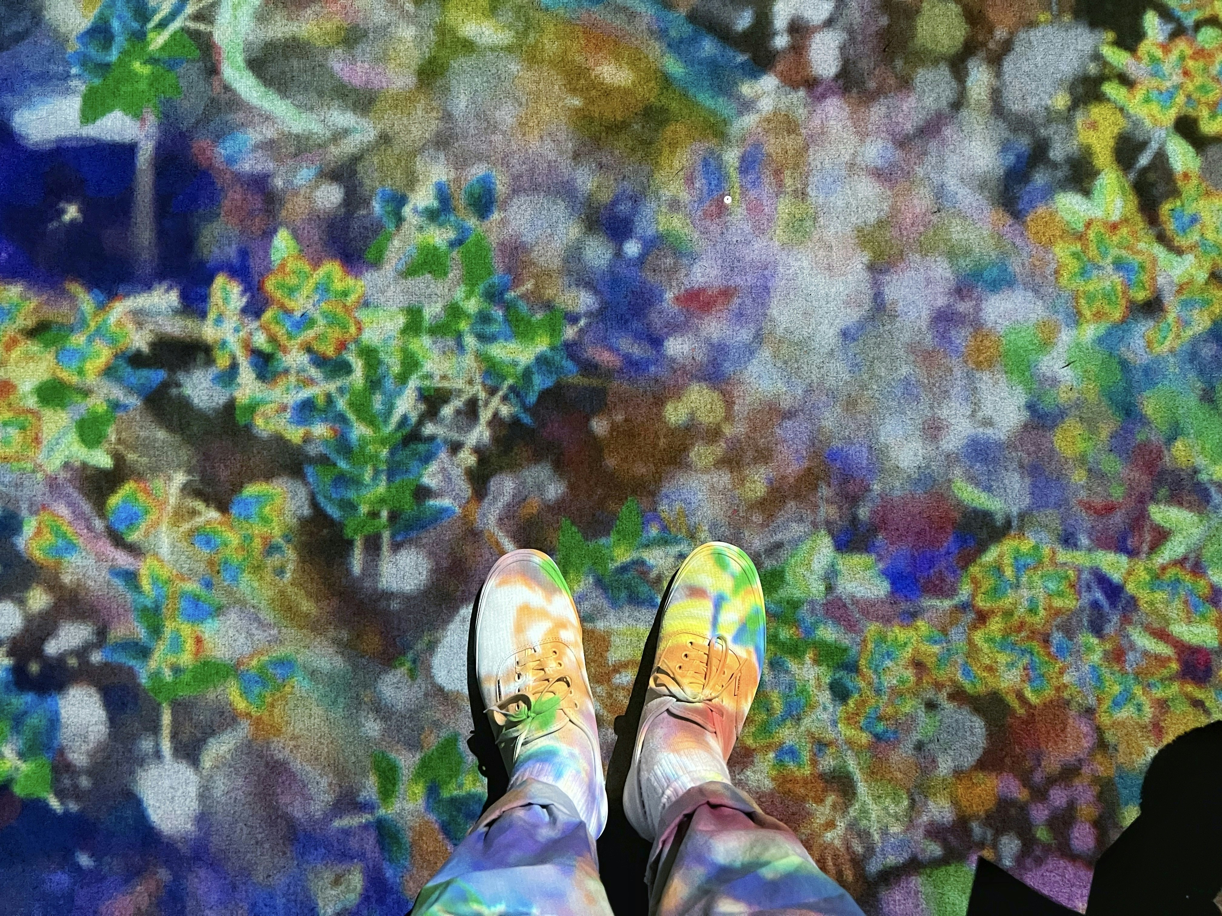 Colorful abstract patterns projected onto the ground, with a pair of feet showcasing vibrant footwear. The scene evokes a playful interaction with the art.