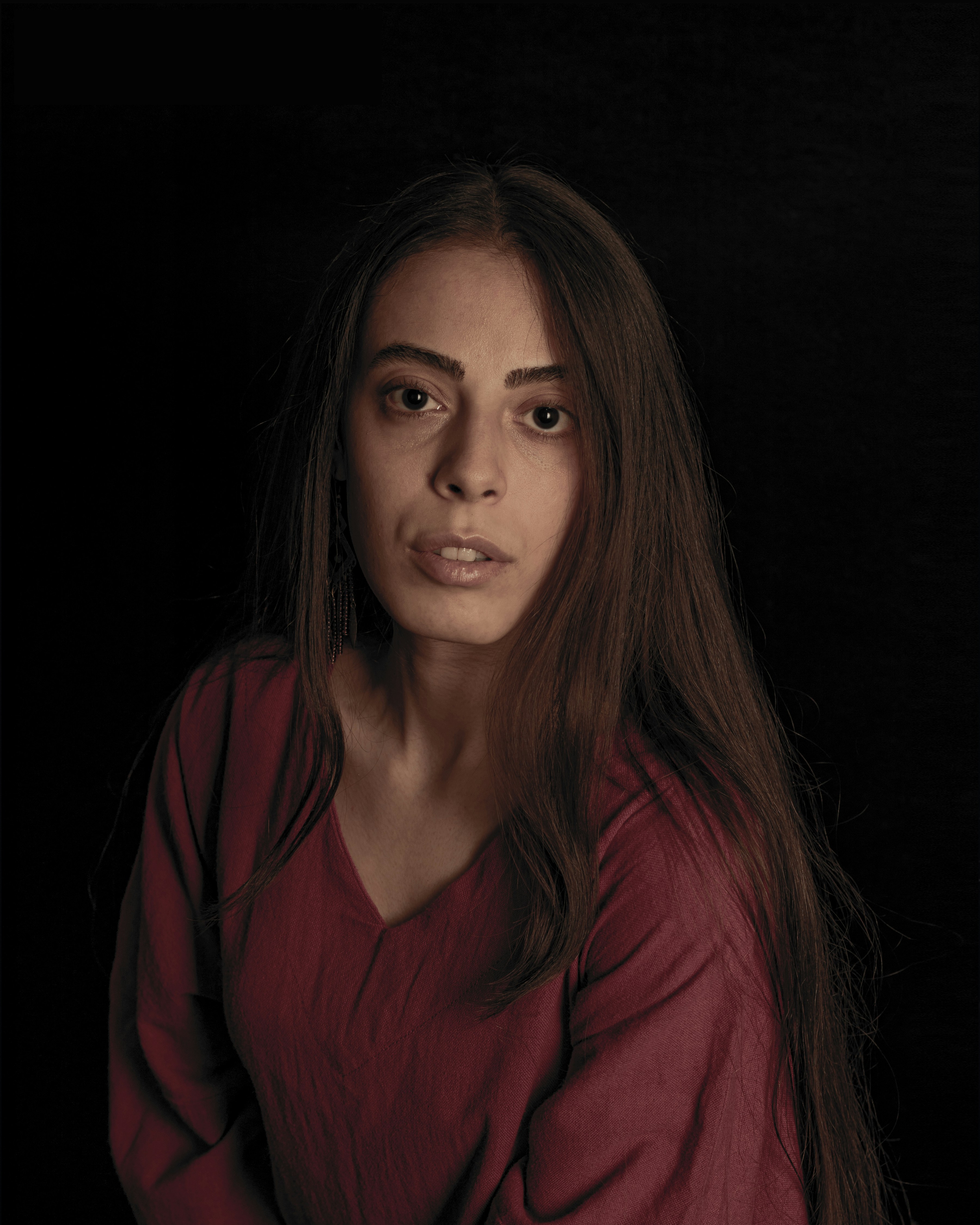A woman with long hair gazes intently, dressed in a deep red garment against a dark background, conveying a sense of introspection.