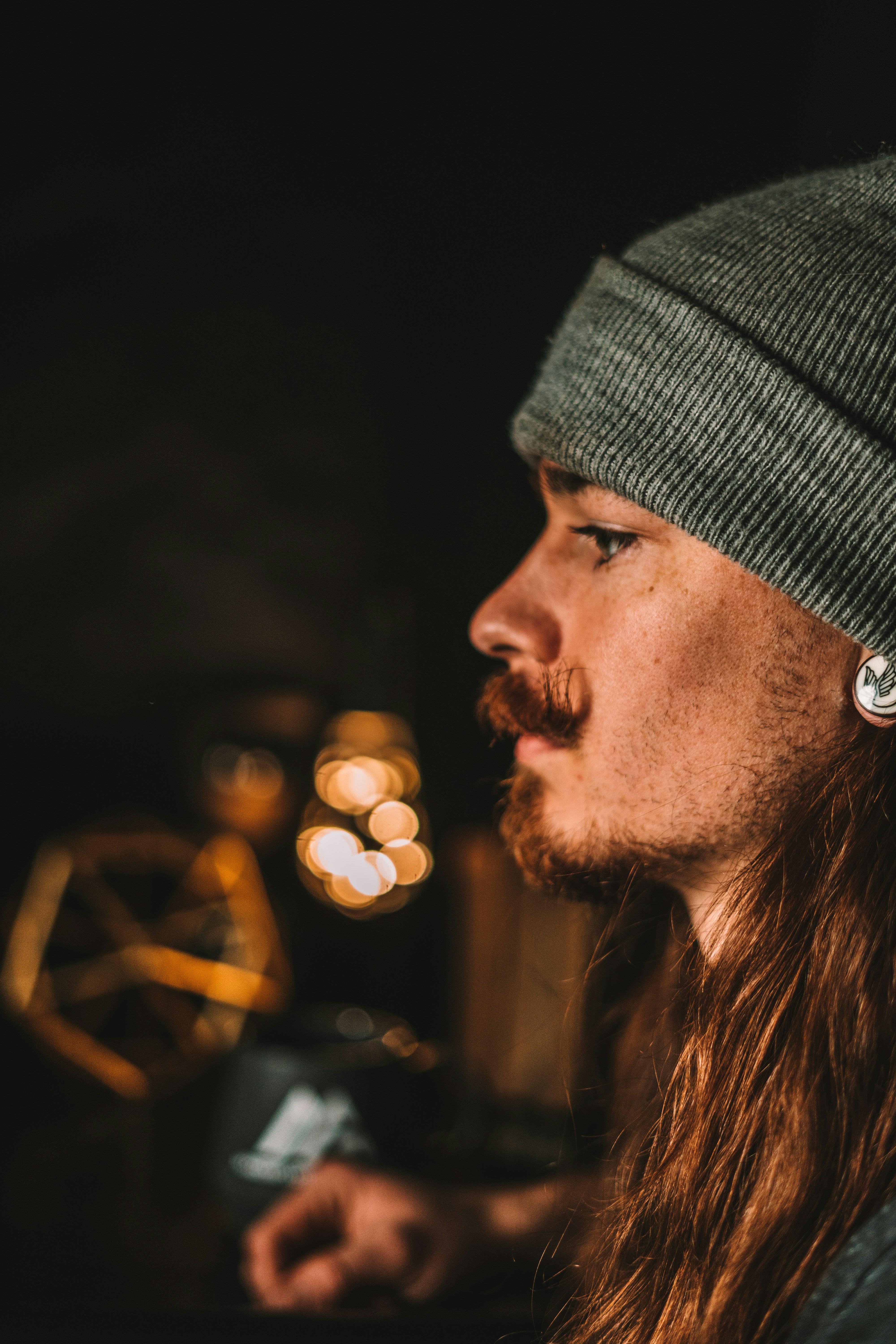 Profile of a man with long hair and a beanie, immersed in thought against a backdrop of warm, glowing lights.