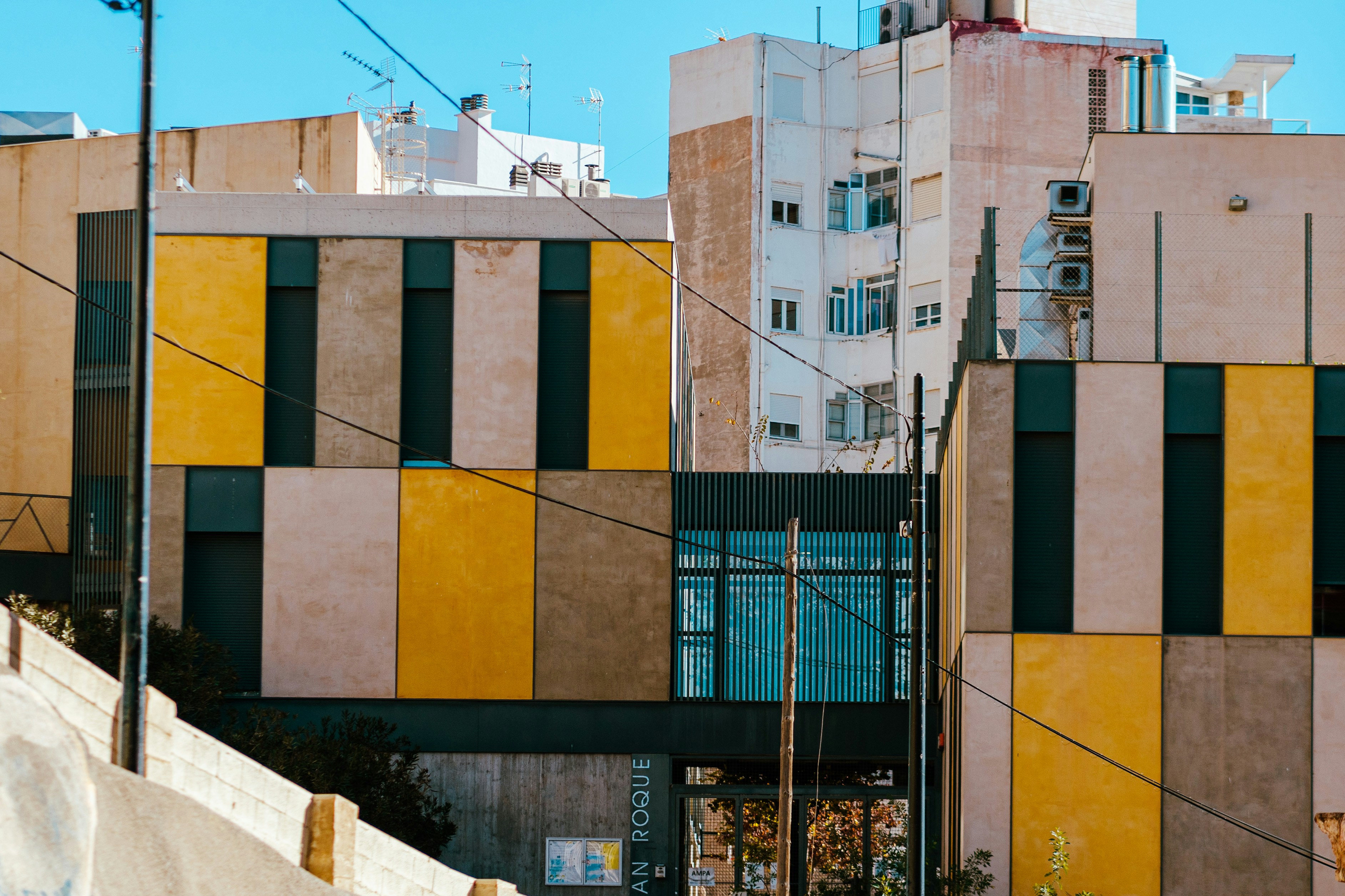 Contemporary building facade showcasing a blend of yellow and muted tones with geometric patterns, set against a backdrop of residential structures.