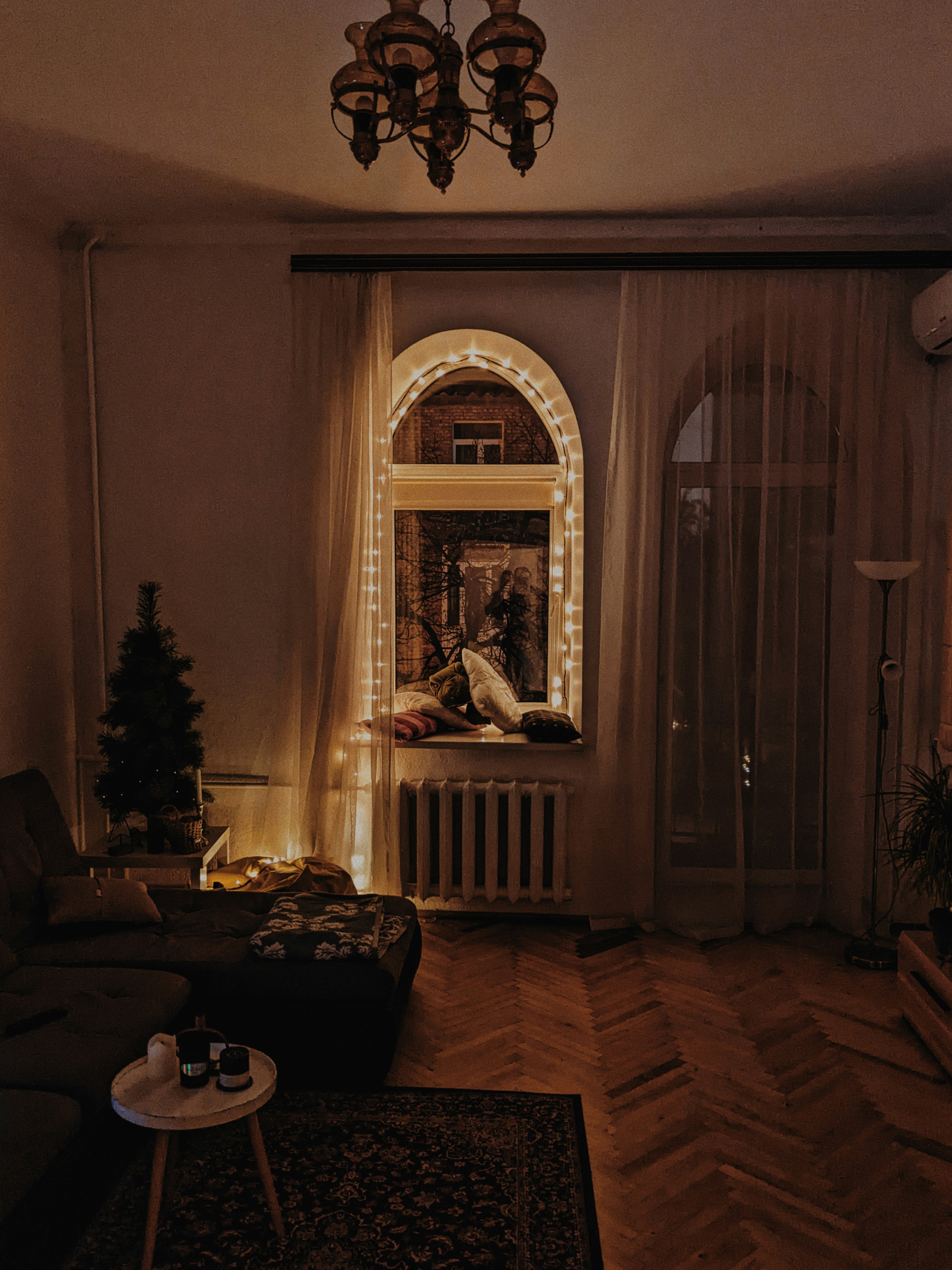 A softly lit living room featuring a window adorned with fairy lights, a small Christmas tree, and a cozy seating area. The ambiance evokes a sense of comfort and tranquility.