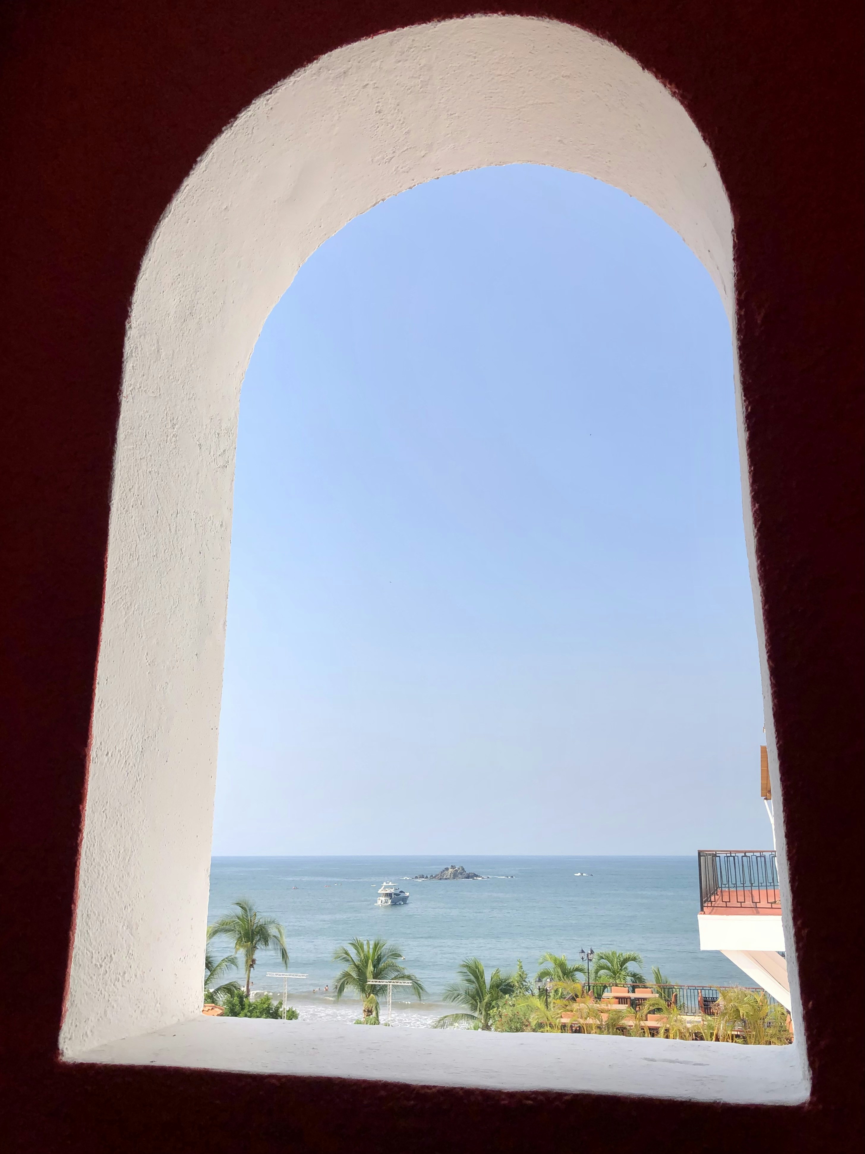 View through an arched window showcasing a tranquil beach scene with boats and palm trees under a clear blue sky.