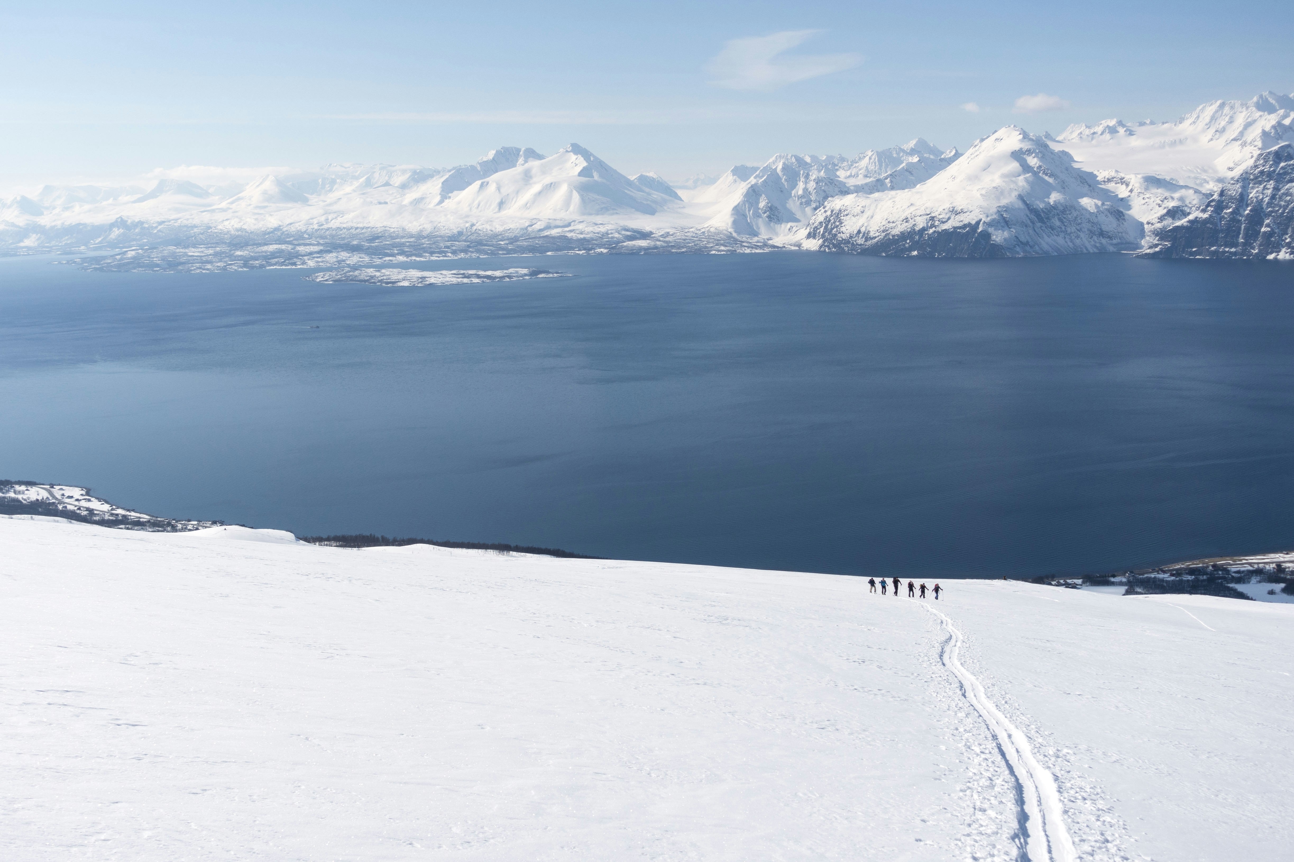 Ski tracks lead across a vast snowy expanse towards the distant Lyngen Alps under a clear blue sky.