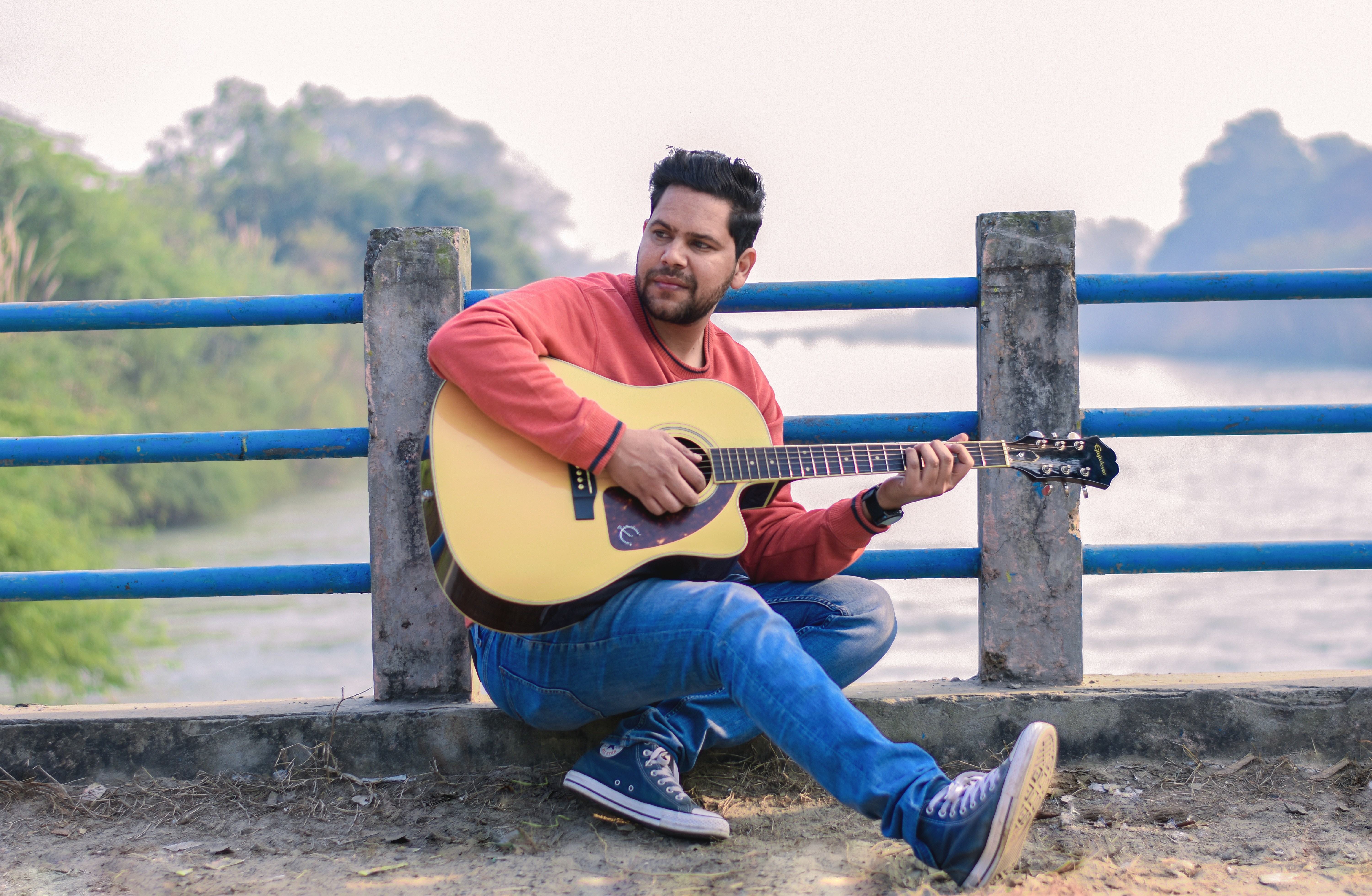 A young man strums an acoustic guitar while seated beside a serene river, framed by blue railings and lush greenery in the background.
