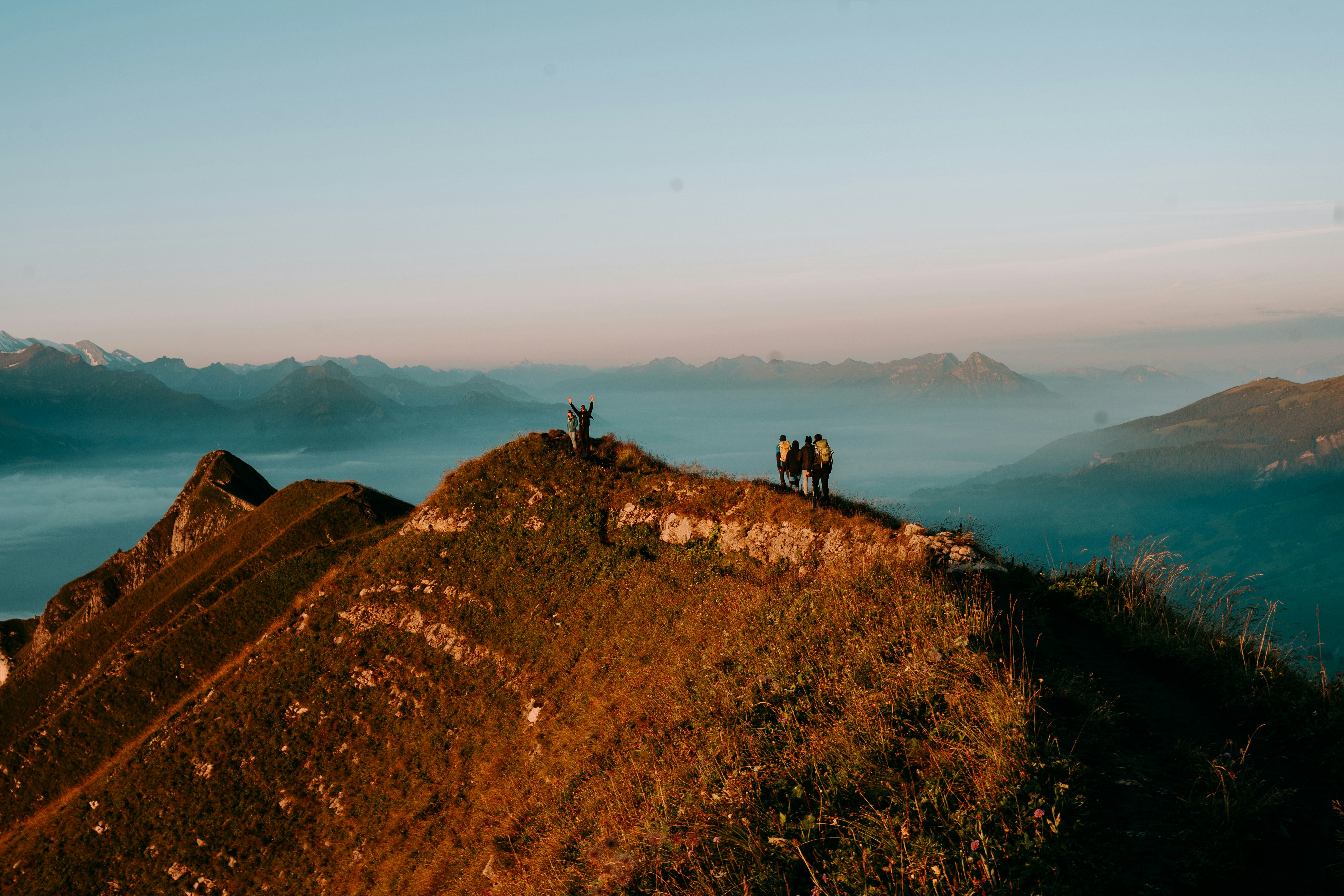 Hikers stand atop a mountain ridge, surrounded by misty valleys and distant peaks at dawn. The scene captures the tranquility and beauty of the alpine landscape.