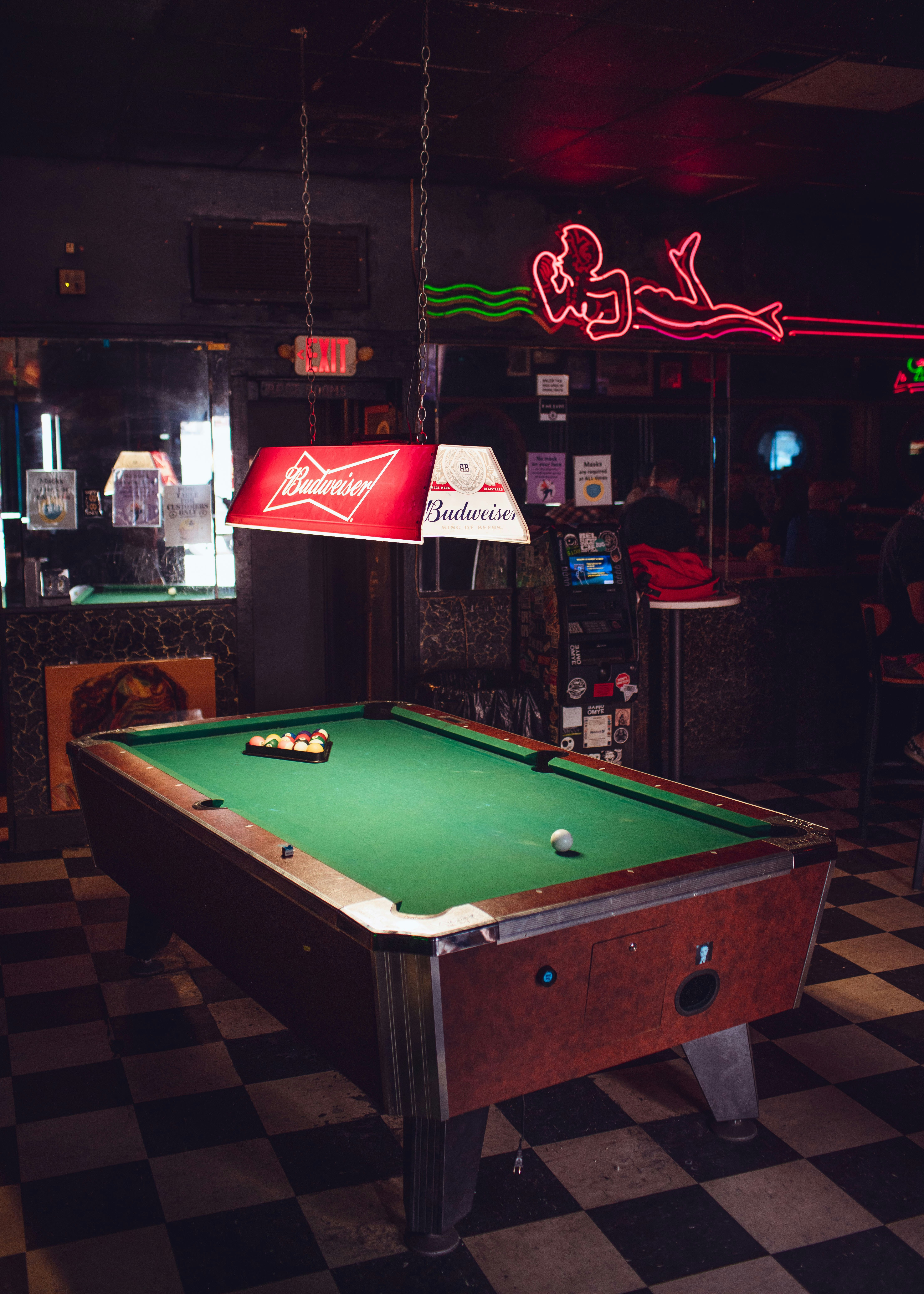 Pool table with balls arranged for a game, illuminated by a Budweiser light fixture in a dimly lit bar setting.