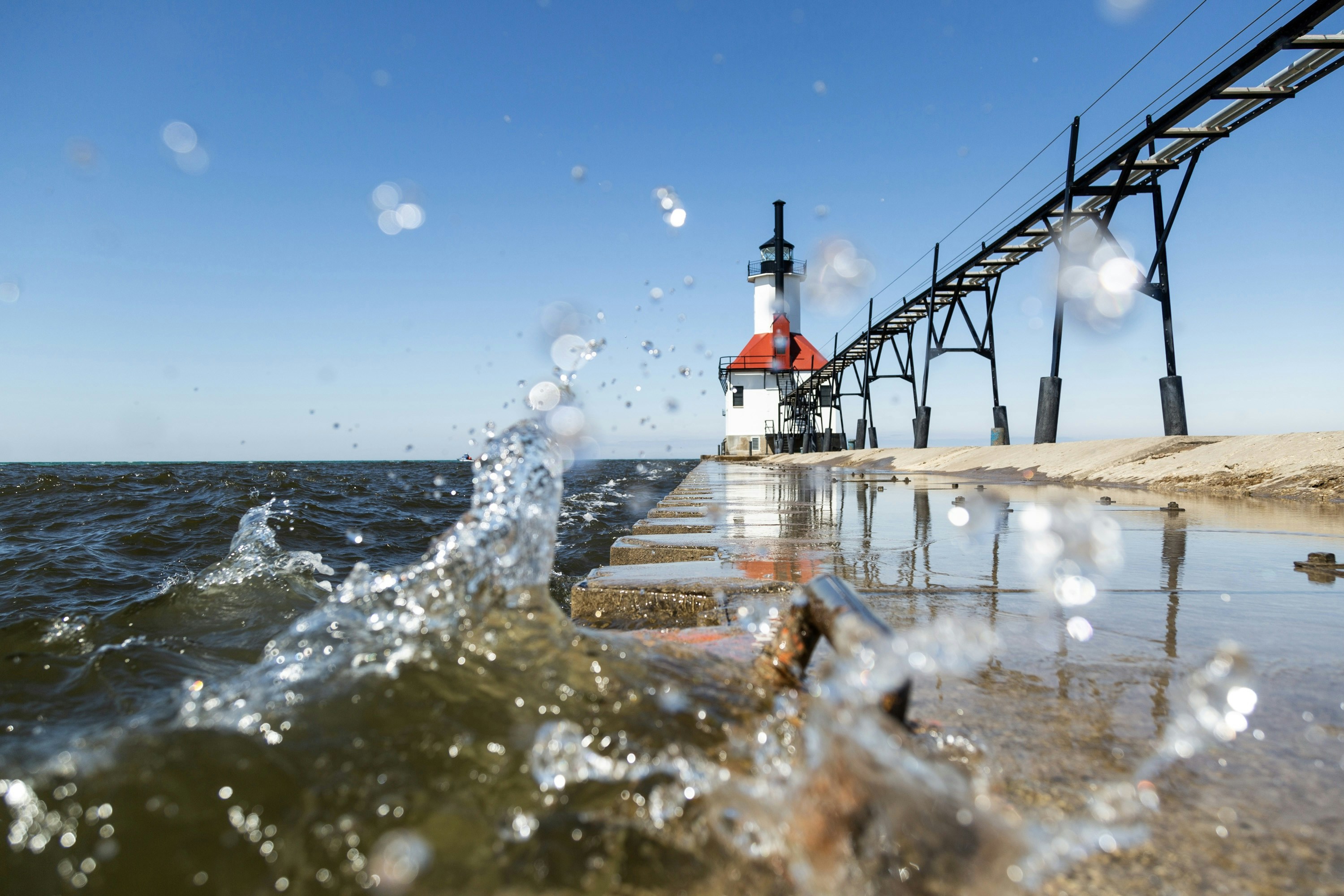 Lighthouse standing tall at the edge of a pier, with splashes of water in the foreground under a clear blue sky.