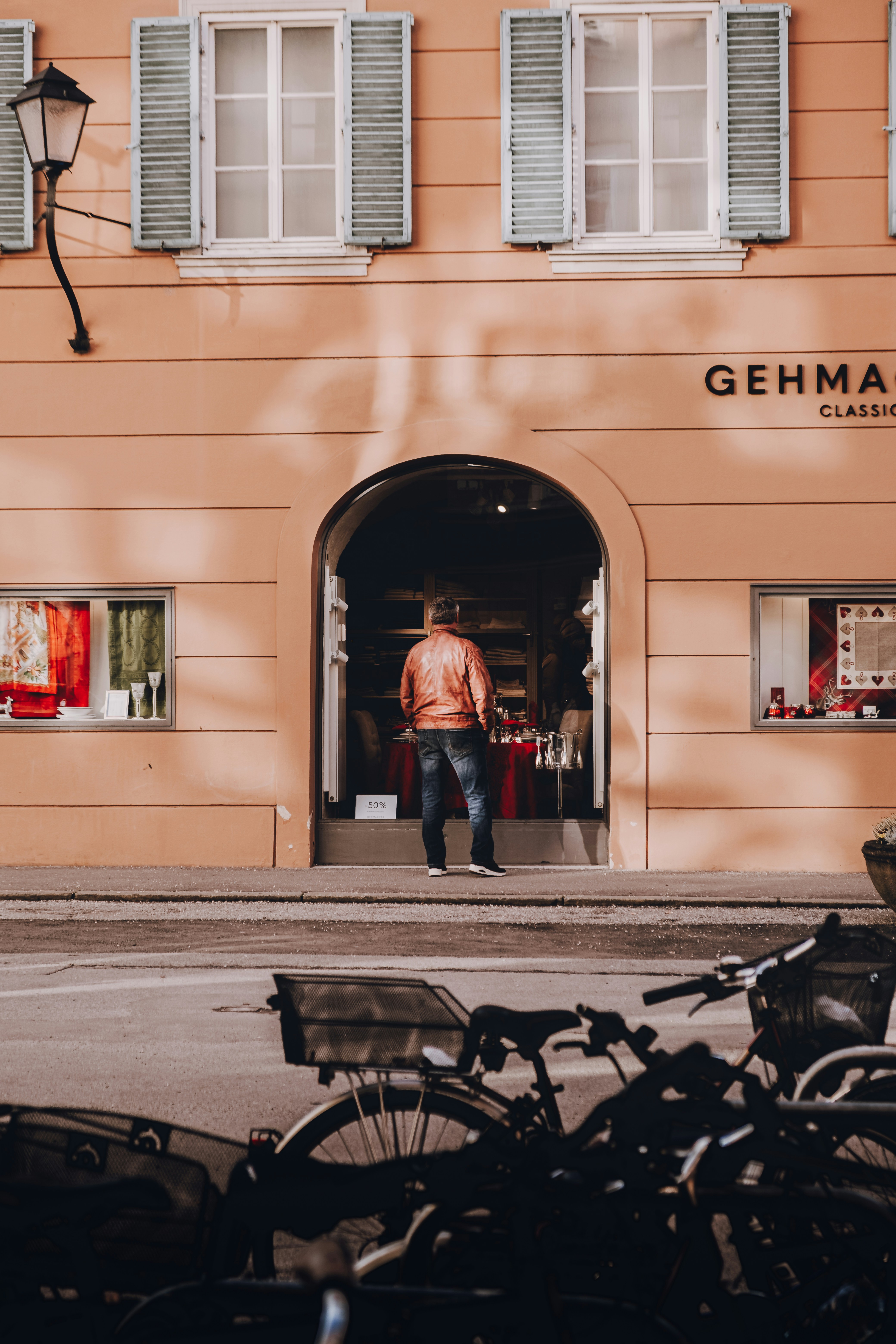 Man in a brown leather jacket stands at the entrance of a charming shop, framed by colorful window displays and a warm peach-colored wall.