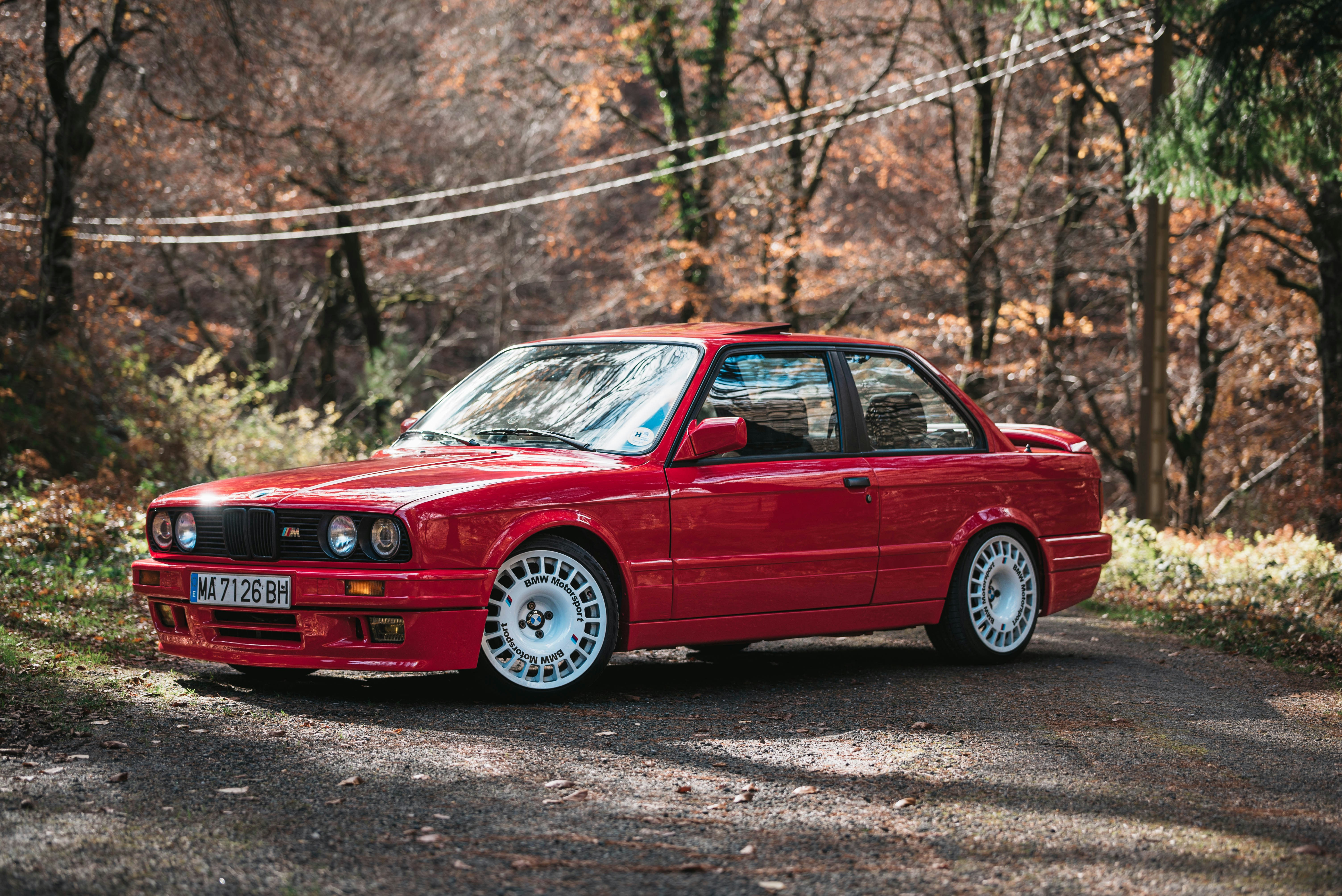 Red vintage car parked on a quiet forest road surrounded by autumn foliage.