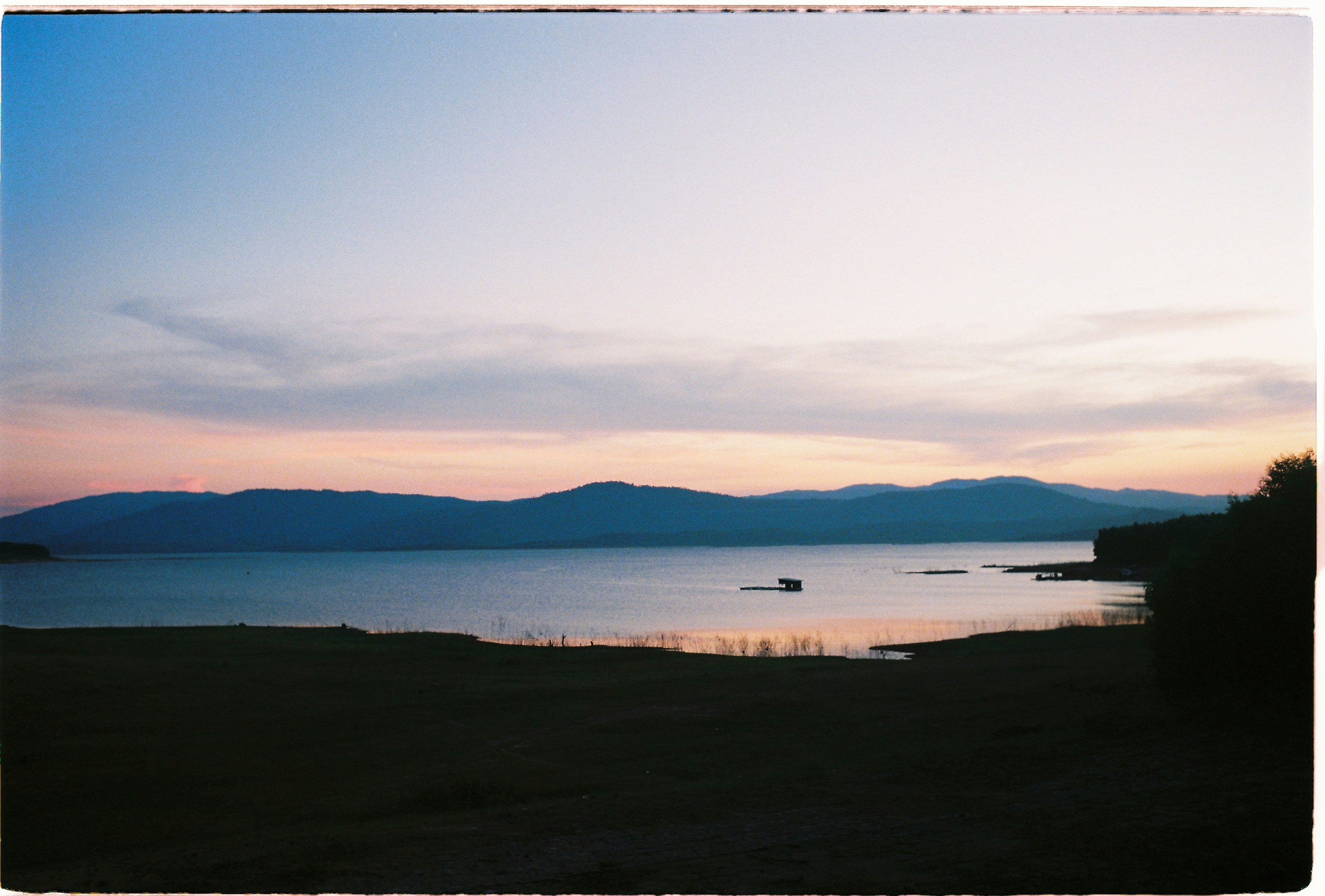 Soft twilight over a lake with distant hills; a calm shoreline and a small boat resting near the water. A serene landscape scene, captured in a single exposure.