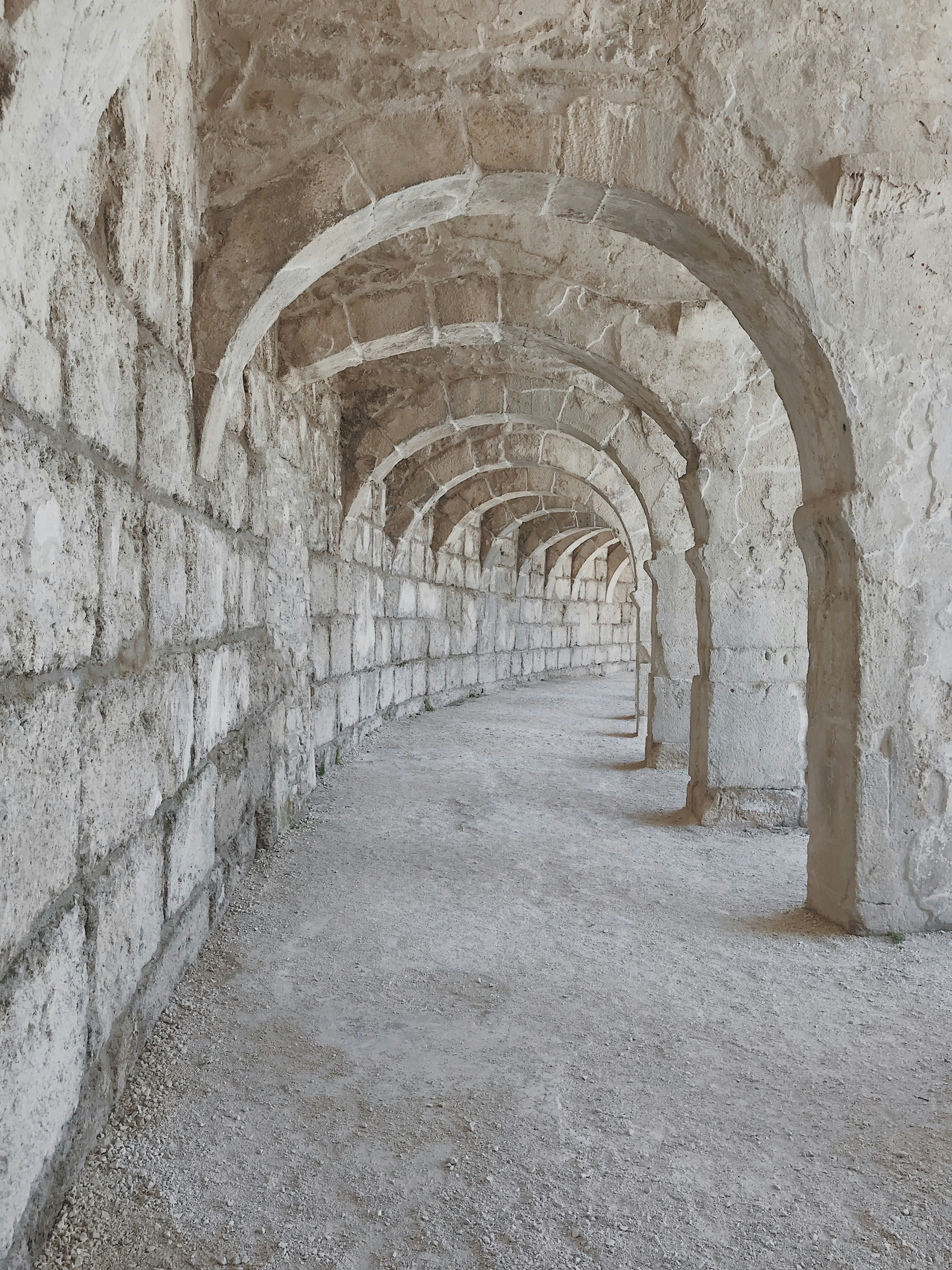 A stone corridor with multiple arches, leading into a softly lit pathway, showcasing the texture of aged walls and gravel floor.