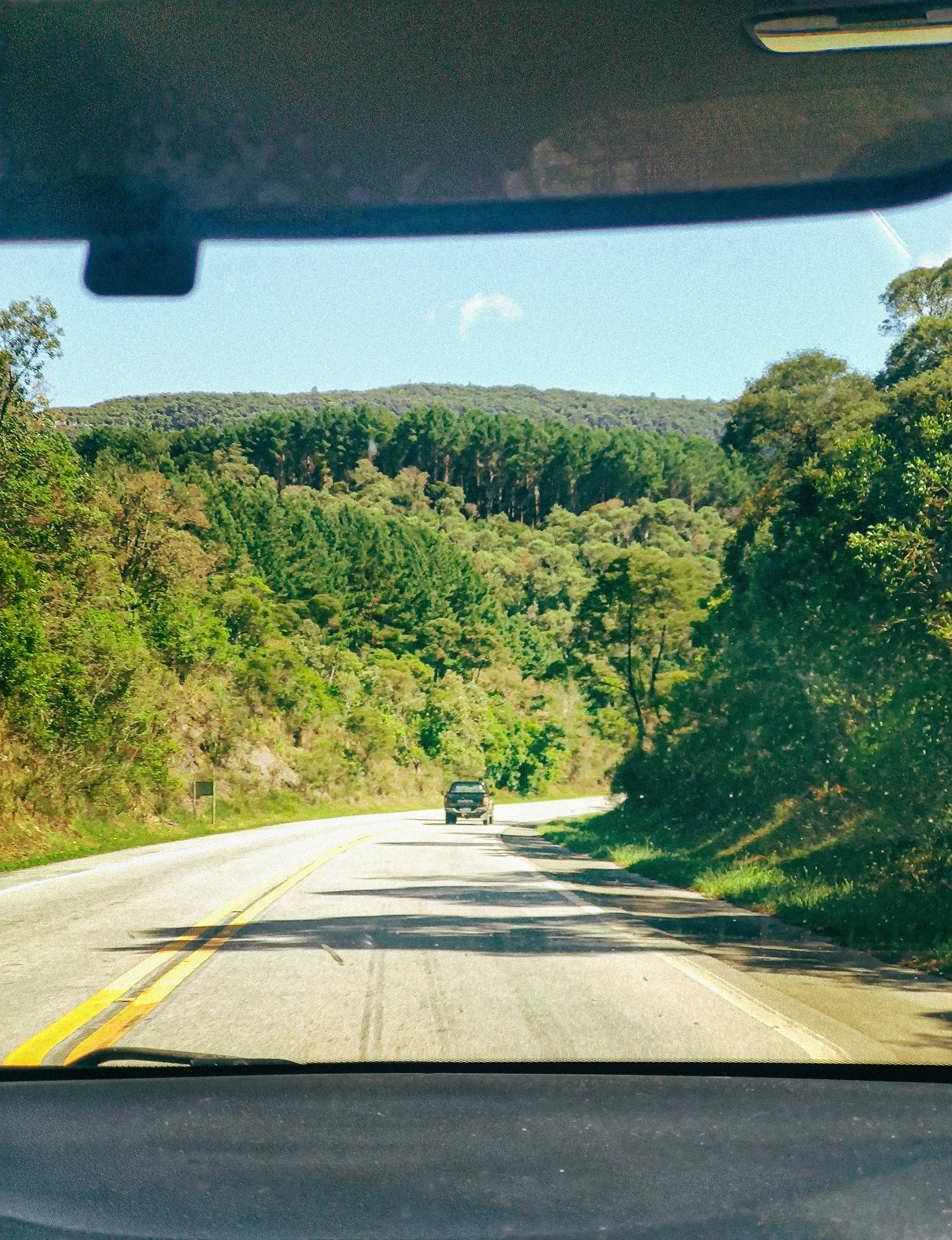 A scenic view from a vehicle showcasing a curving road flanked by lush greenery and distant hills under a clear blue sky.