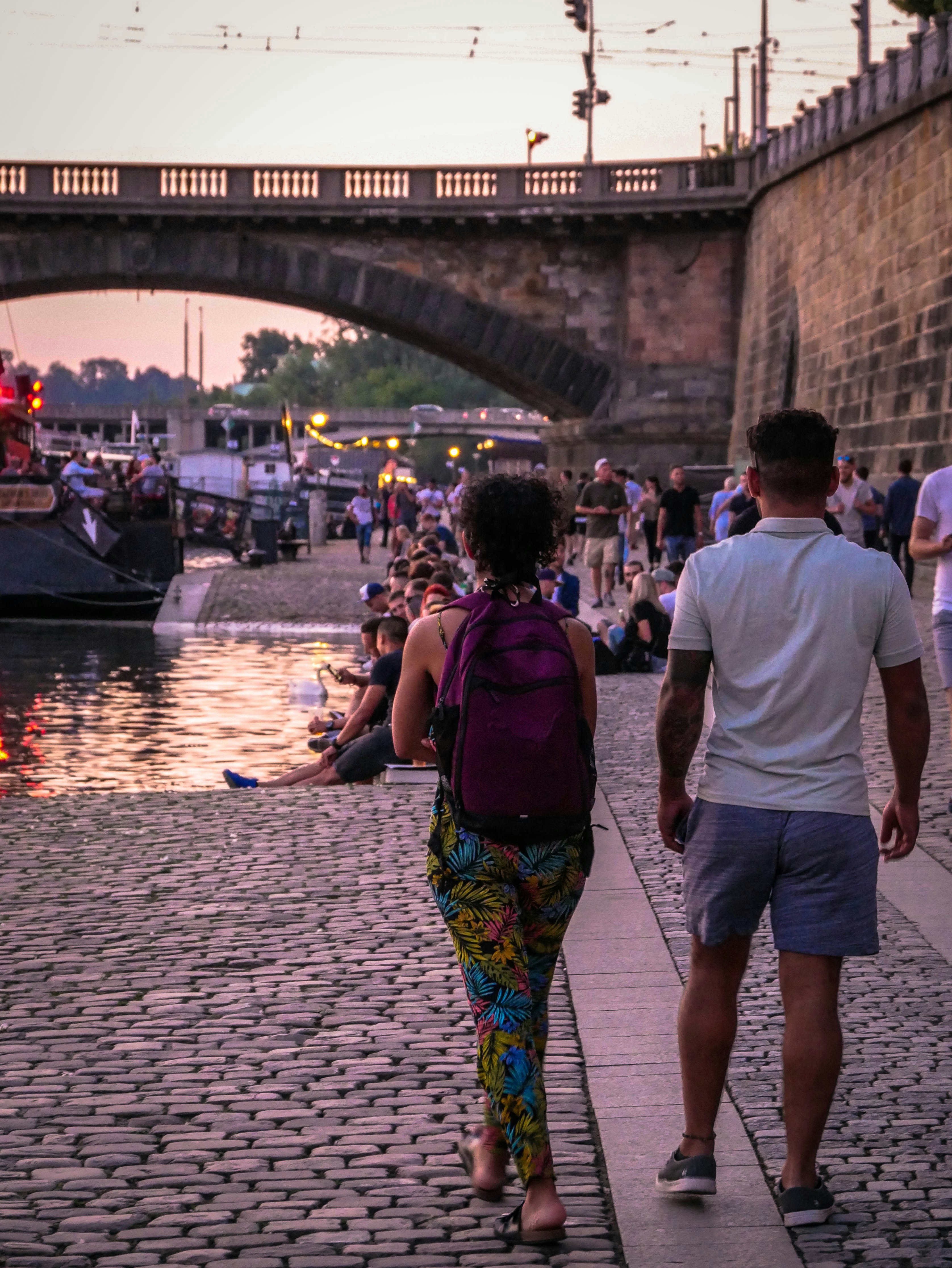 Riverside Evening: A Purple-Backpacked Figure Leads the Cobblestone Promenade