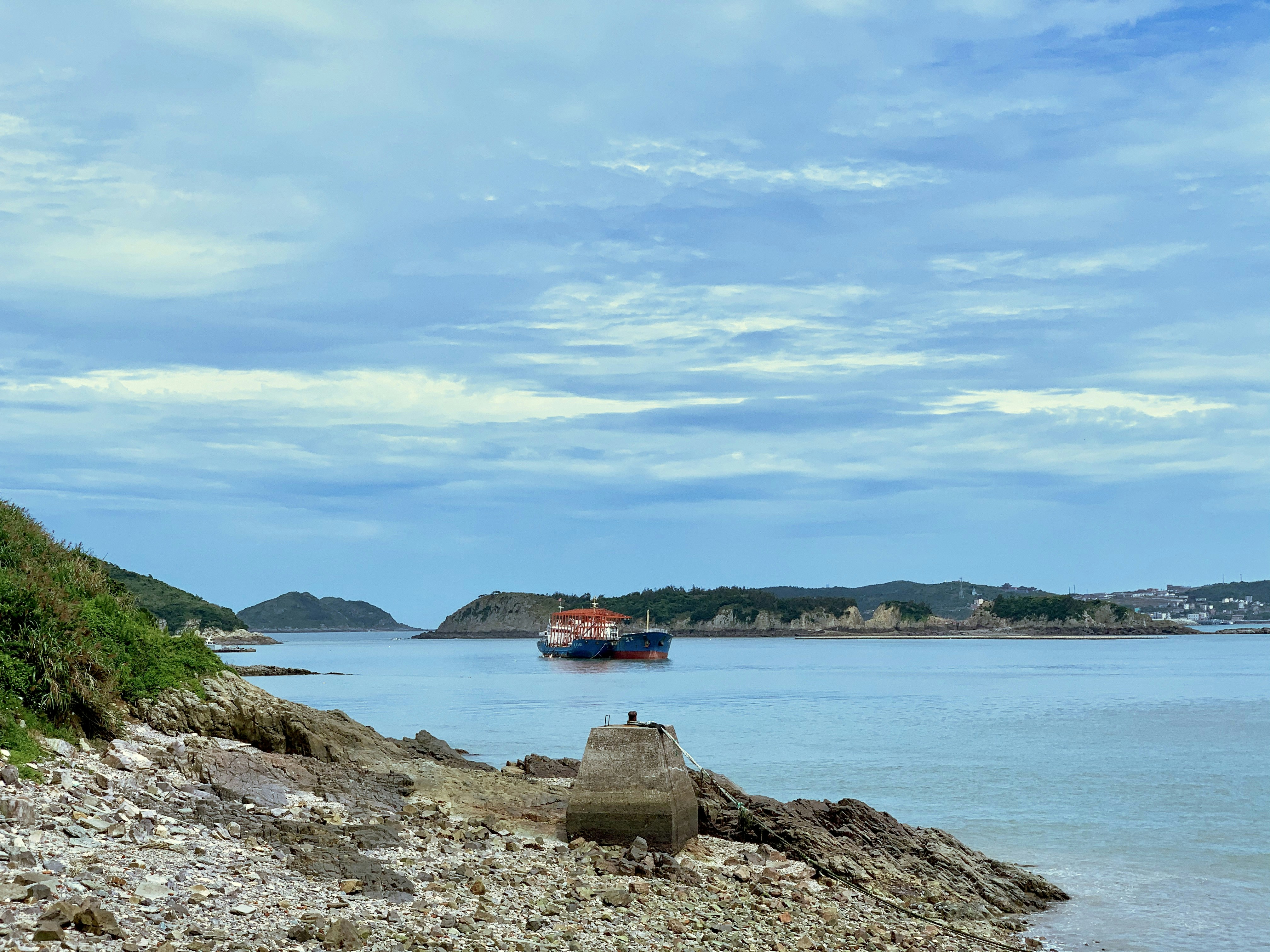 A cargo ship anchored in calm waters, framed by rocky shores and distant hills under a cloudy sky.