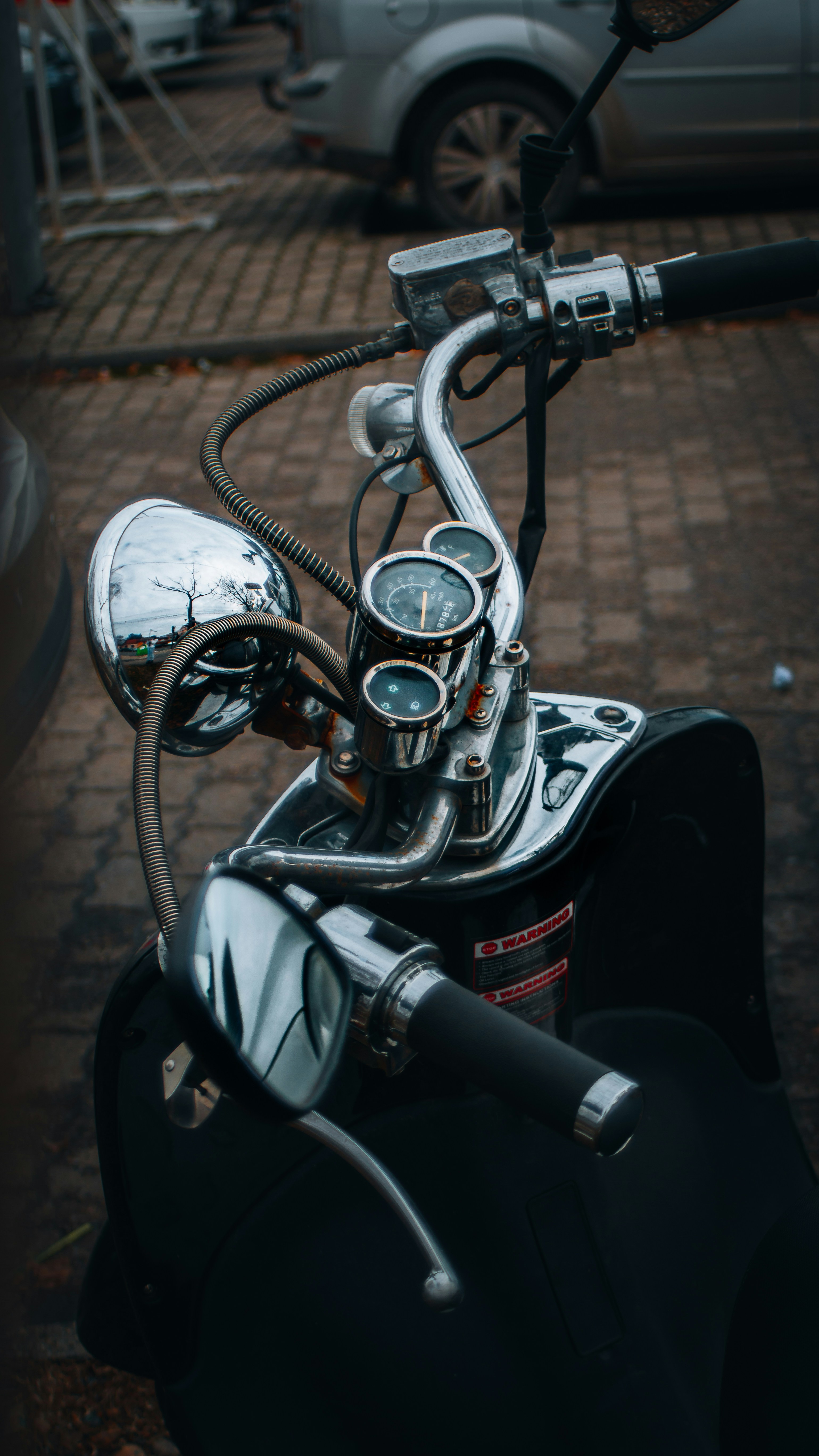 Close-up of a scooter's handlebars featuring chrome details and gauges, reflecting the surroundings. The focus highlights the intricate design and craftsmanship.