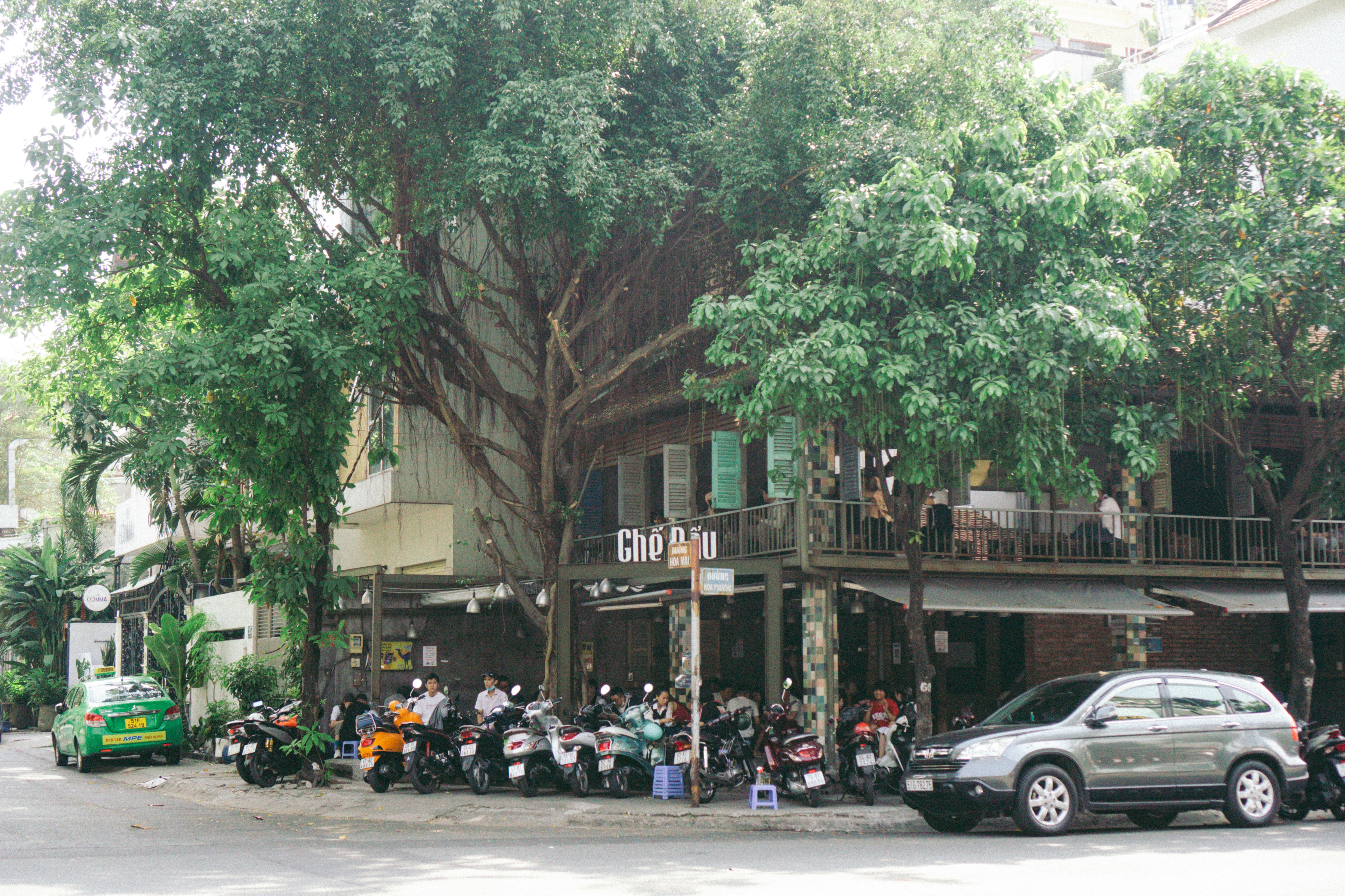 A bustling street scene featuring a vibrant café surrounded by lush trees and parked motorcycles. Patrons enjoy their time in a shaded outdoor setting.