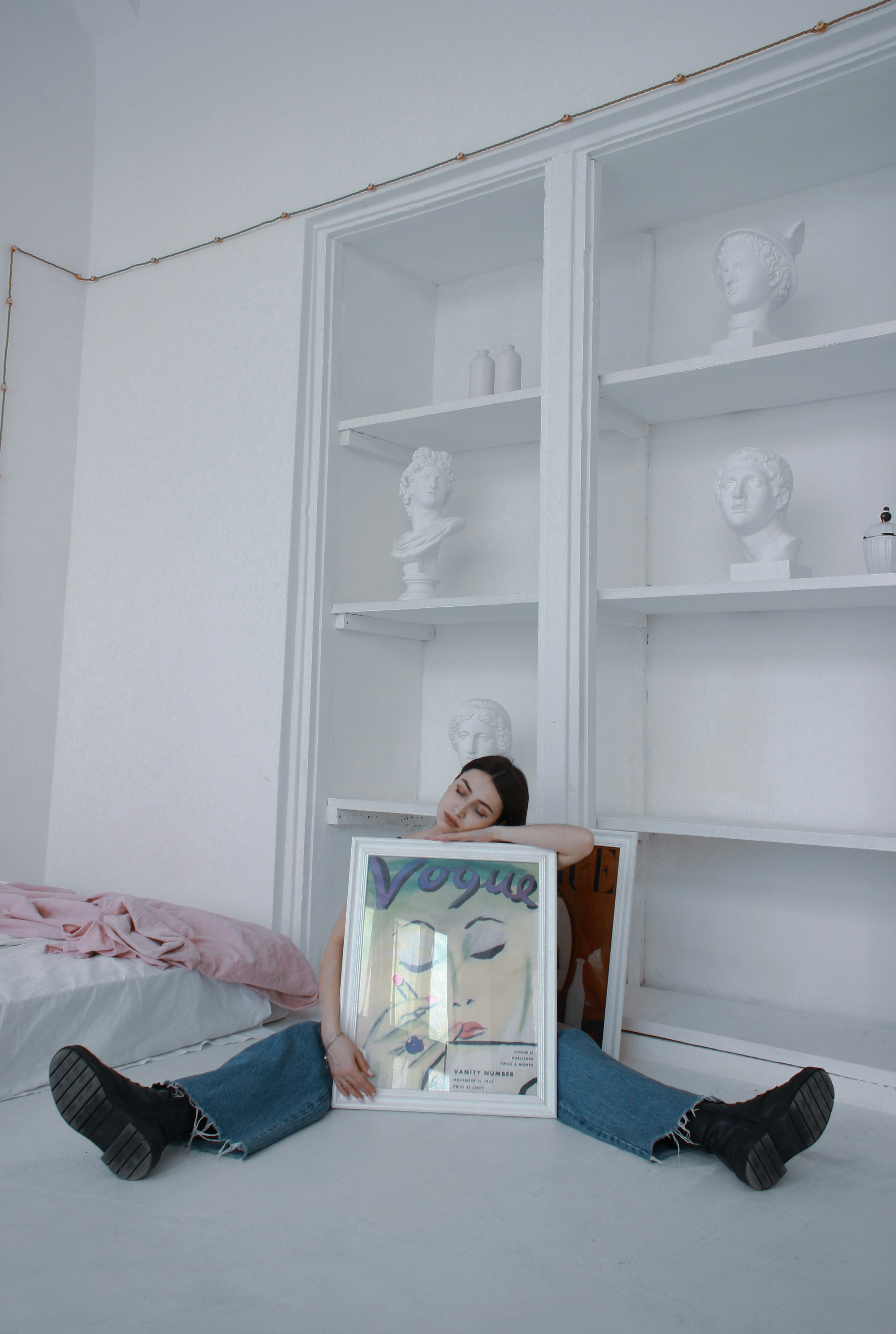 Person seated on a white floor cradling a framed artwork, surrounded by empty white shelves with sculpted busts.