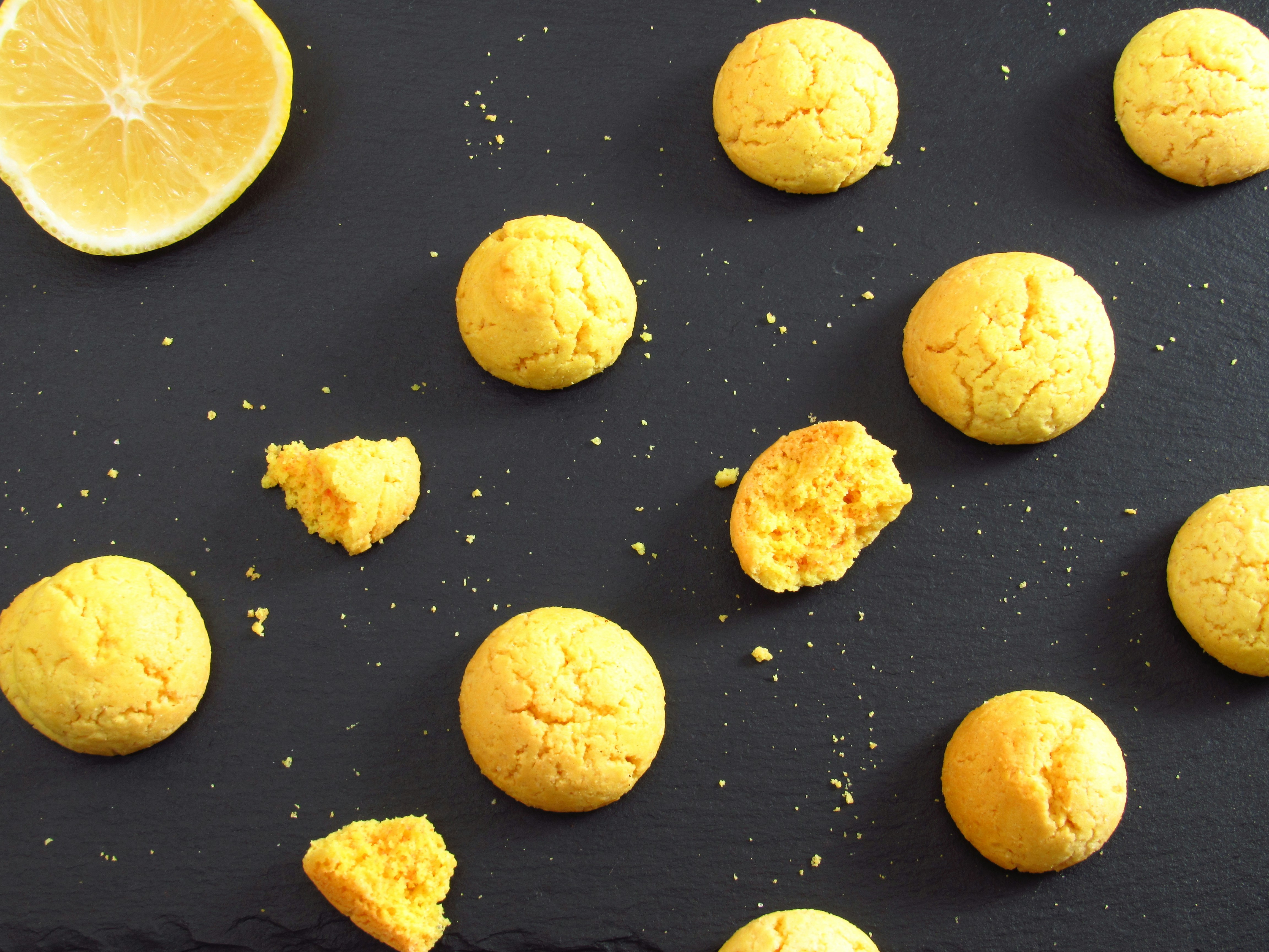 Golden lemon-flavored treats scattered on a dark slate surface, with a fresh lemon slice nearby. Crumbled cookies add texture and contrast.
