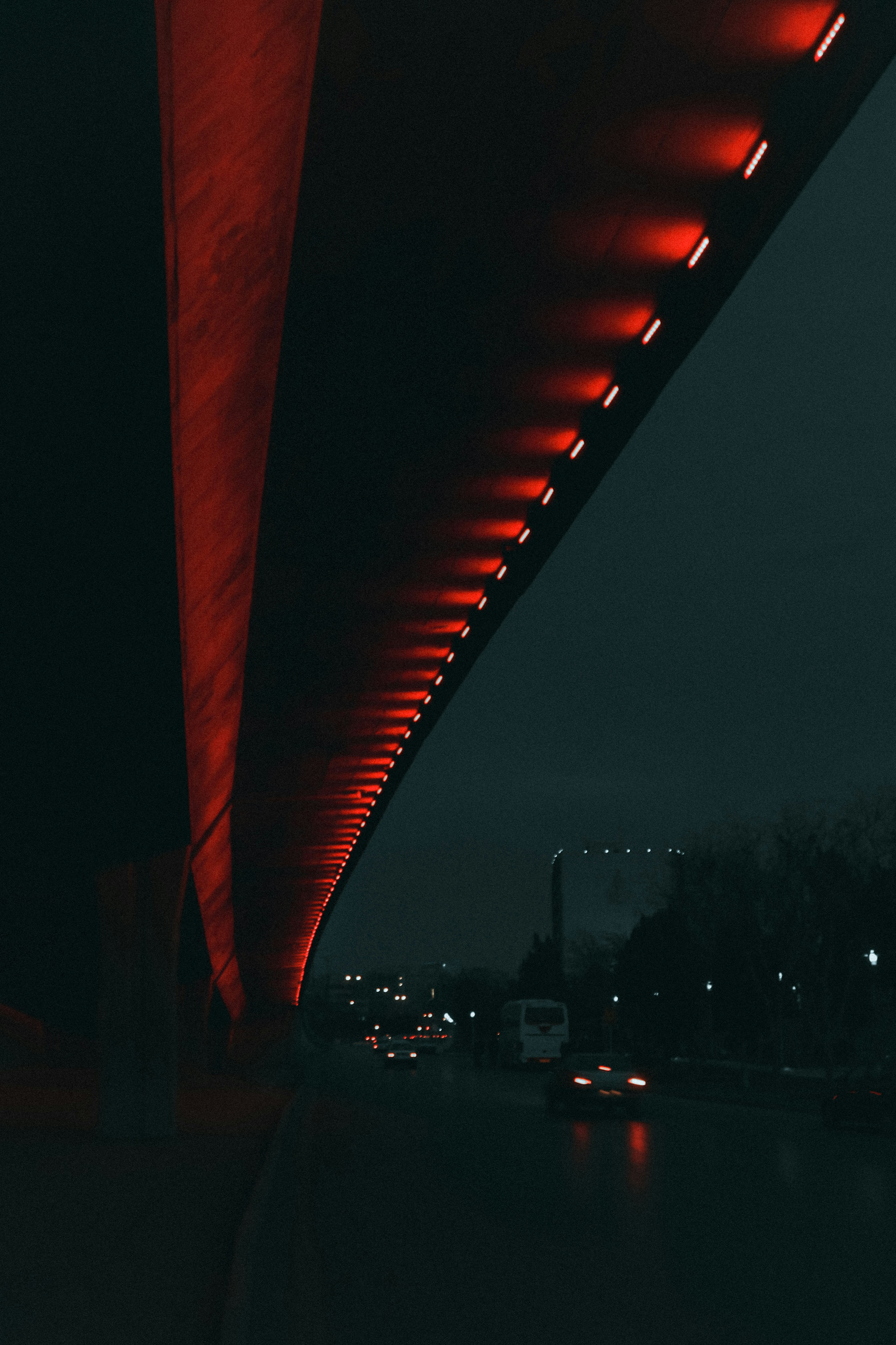 Illuminated underpass with striking red lights casting shadows on the roadway below. Vehicles traverse the darkened scene.