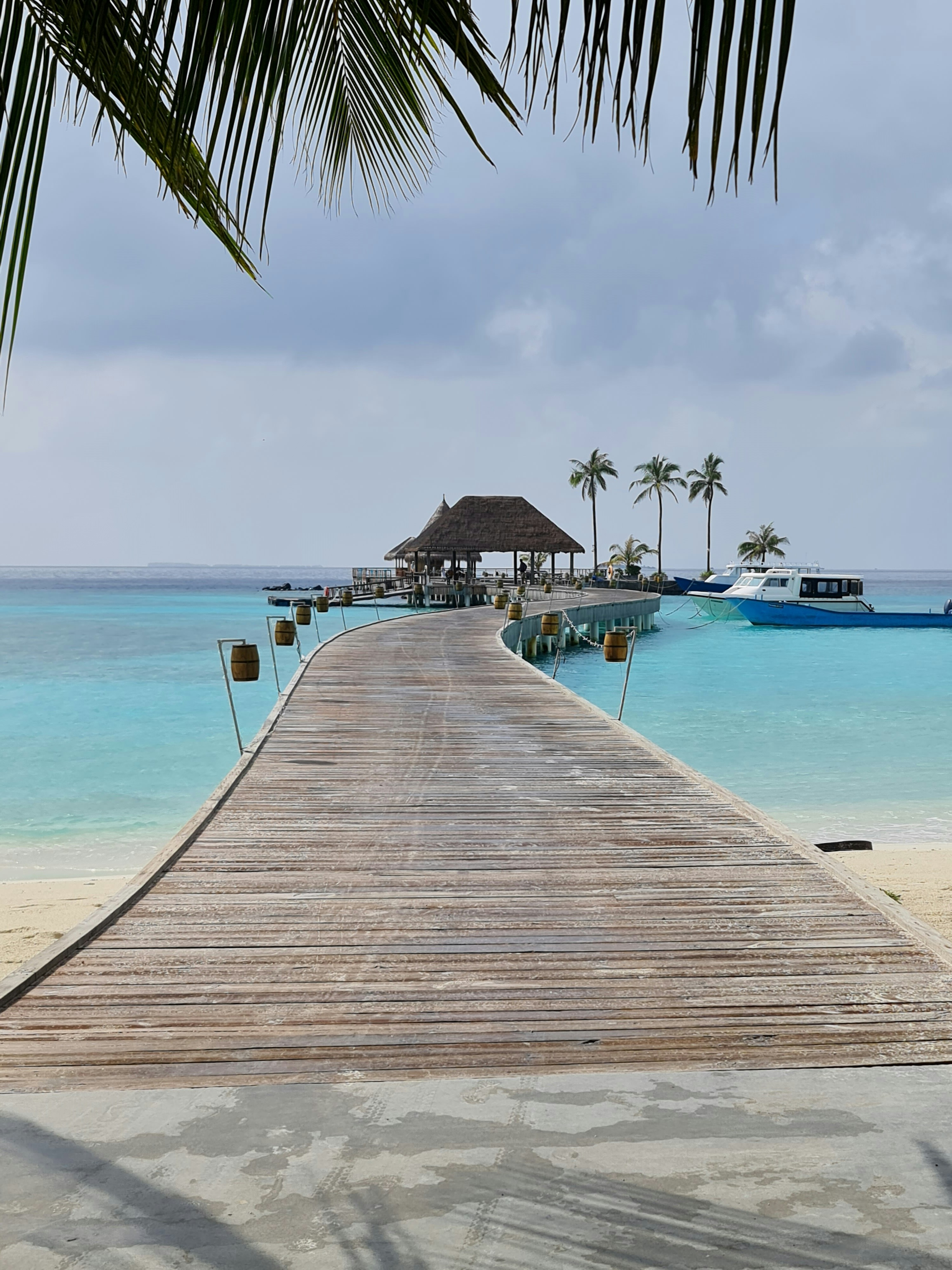 A wooden boardwalk extends through turquoise water toward a thatched-roof pavilion on a small island. Palm trees line the horizon with a docked boat nearby.