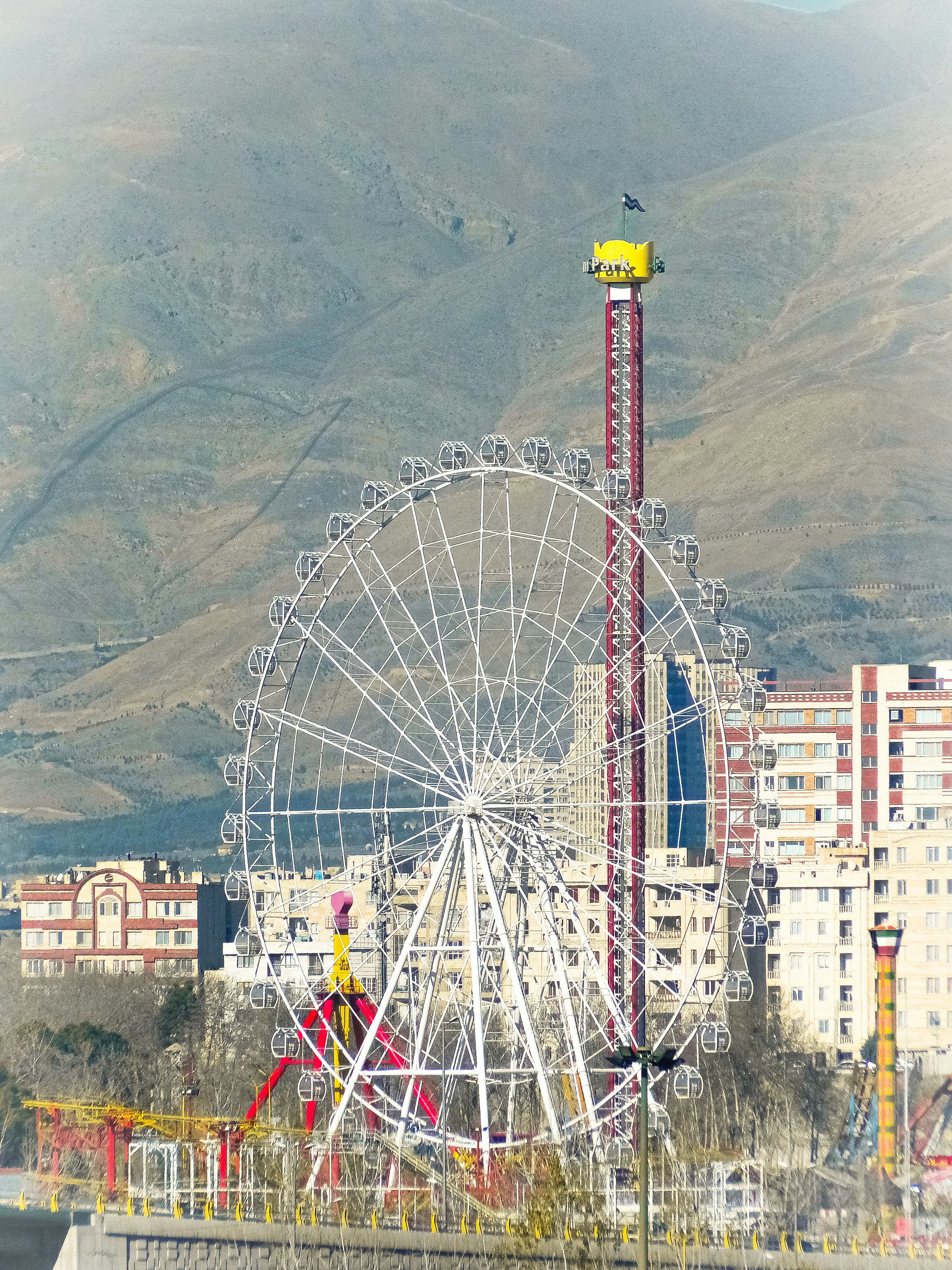 A vibrant ferris wheel and amusement ride stand against a backdrop of majestic mountains and urban architecture. The scene captures the essence of fun and adventure.