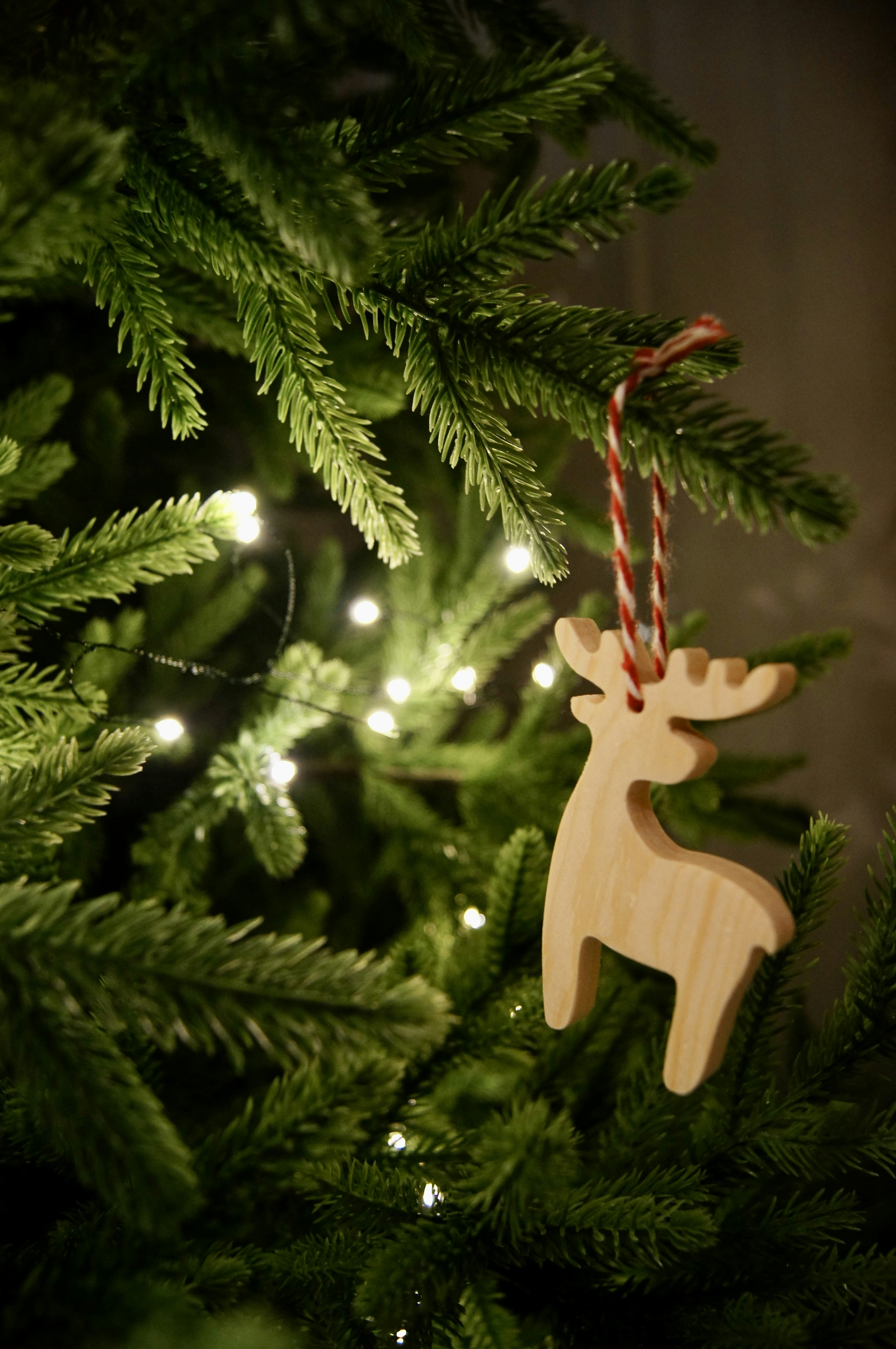 Close-up of a wooden reindeer ornament hanging from a lush Christmas tree with soft bokeh lights in the background. A warm, festive detail shot emphasizing handmade decor.