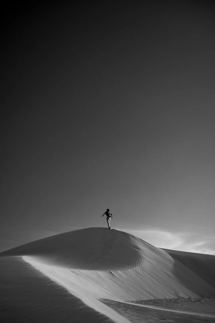 A lone person stands on the crest of a sand dune under a vast dark sky, with sweeping ripples and strong light-shadow contrast. Black-and-white composition emphasizes scale and isolation.