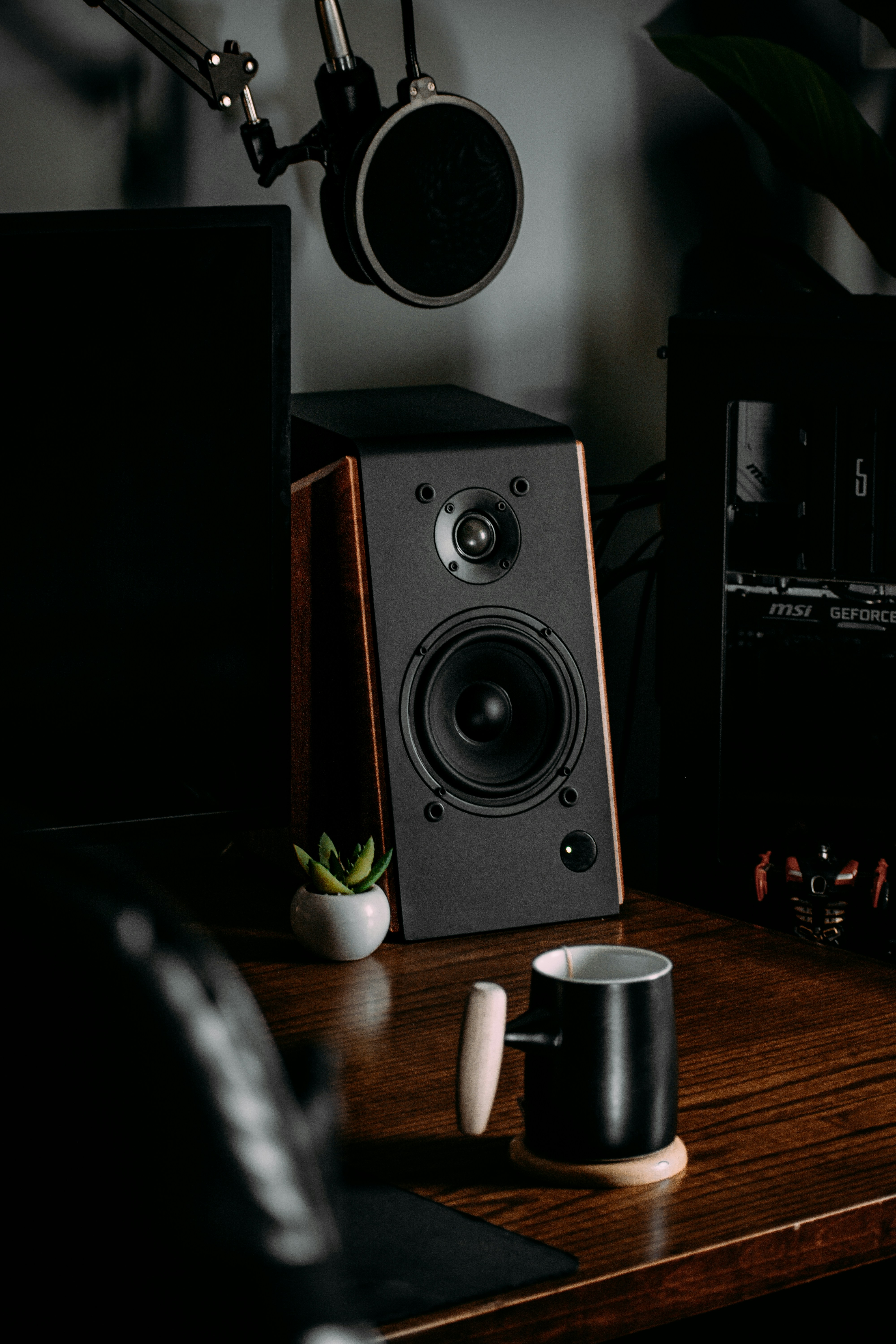 A sleek audio speaker and a steaming mug sit on a wooden desk, accompanied by a small plant, creating a cozy workspace atmosphere.