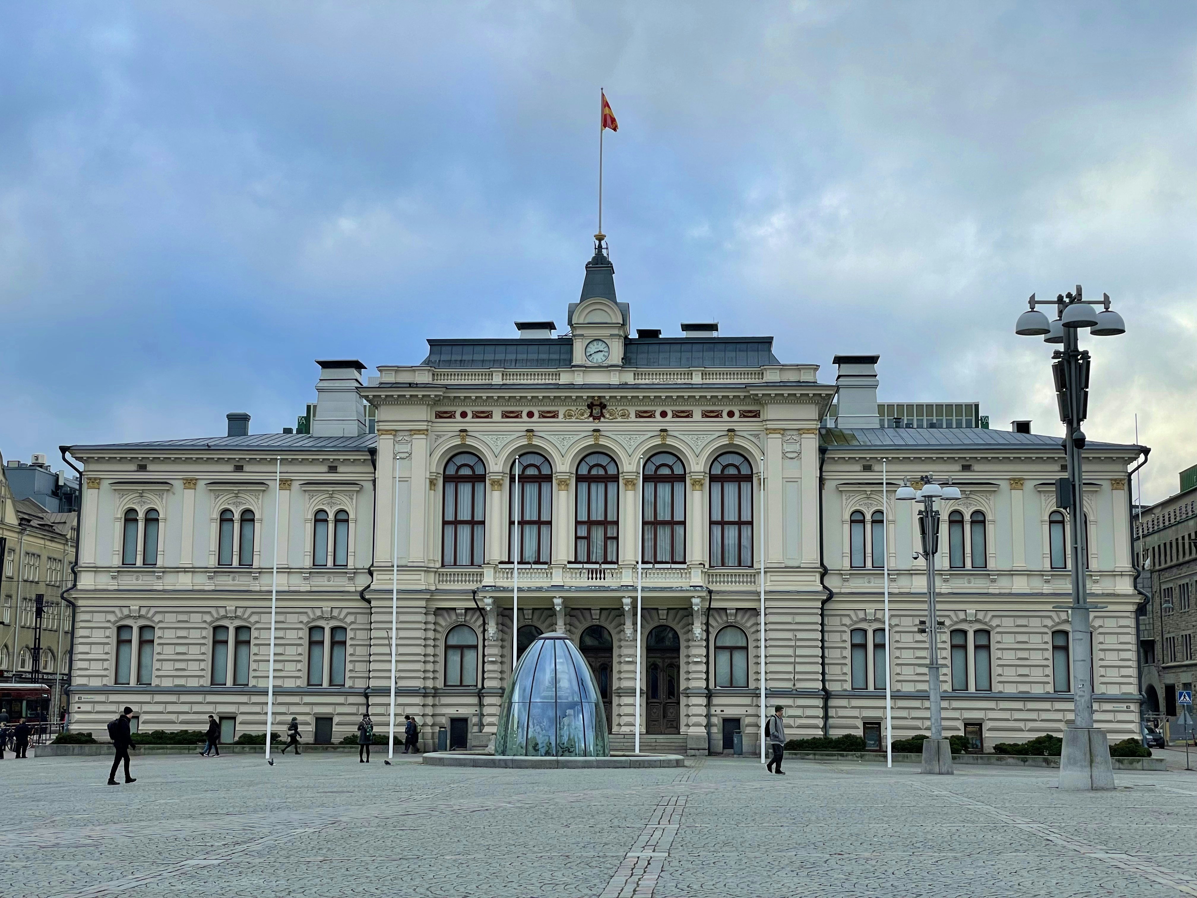 Historic building featuring classical architecture with an artistic installation in the foreground, set against a cloudy sky.