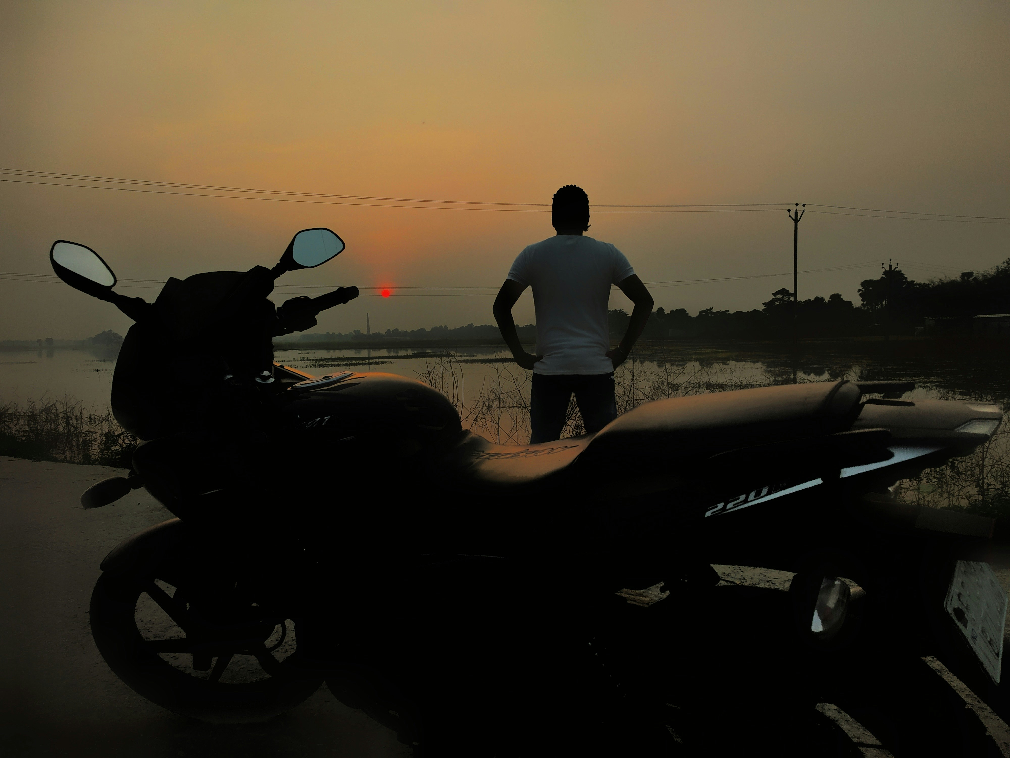 Silhouette of a rider standing beside a motorcycle along a water's edge as the sun dips below the horizon.