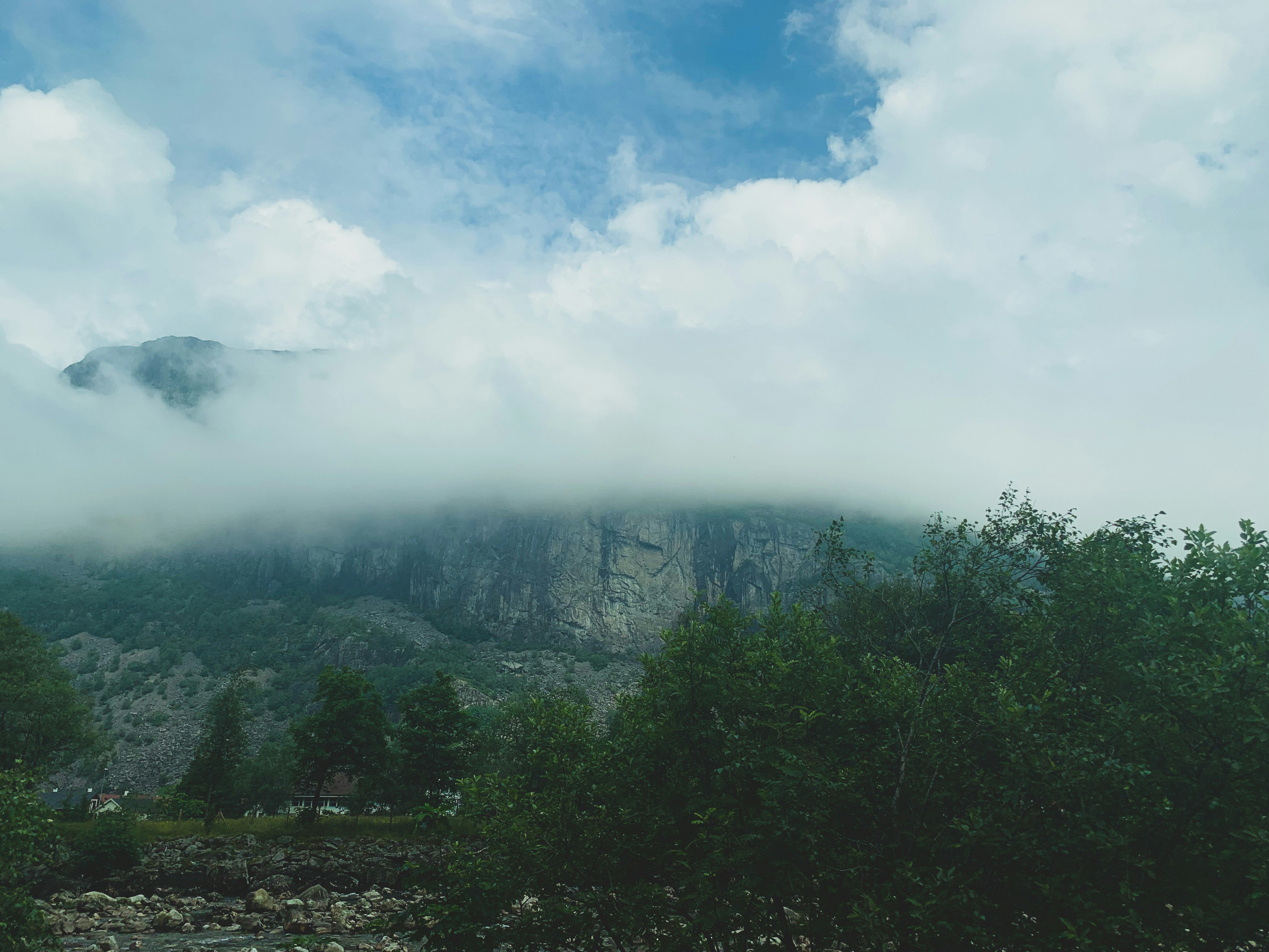 Low-hanging clouds shroud the rugged mountain landscape, partially revealing its towering cliffs and lush greenery below.