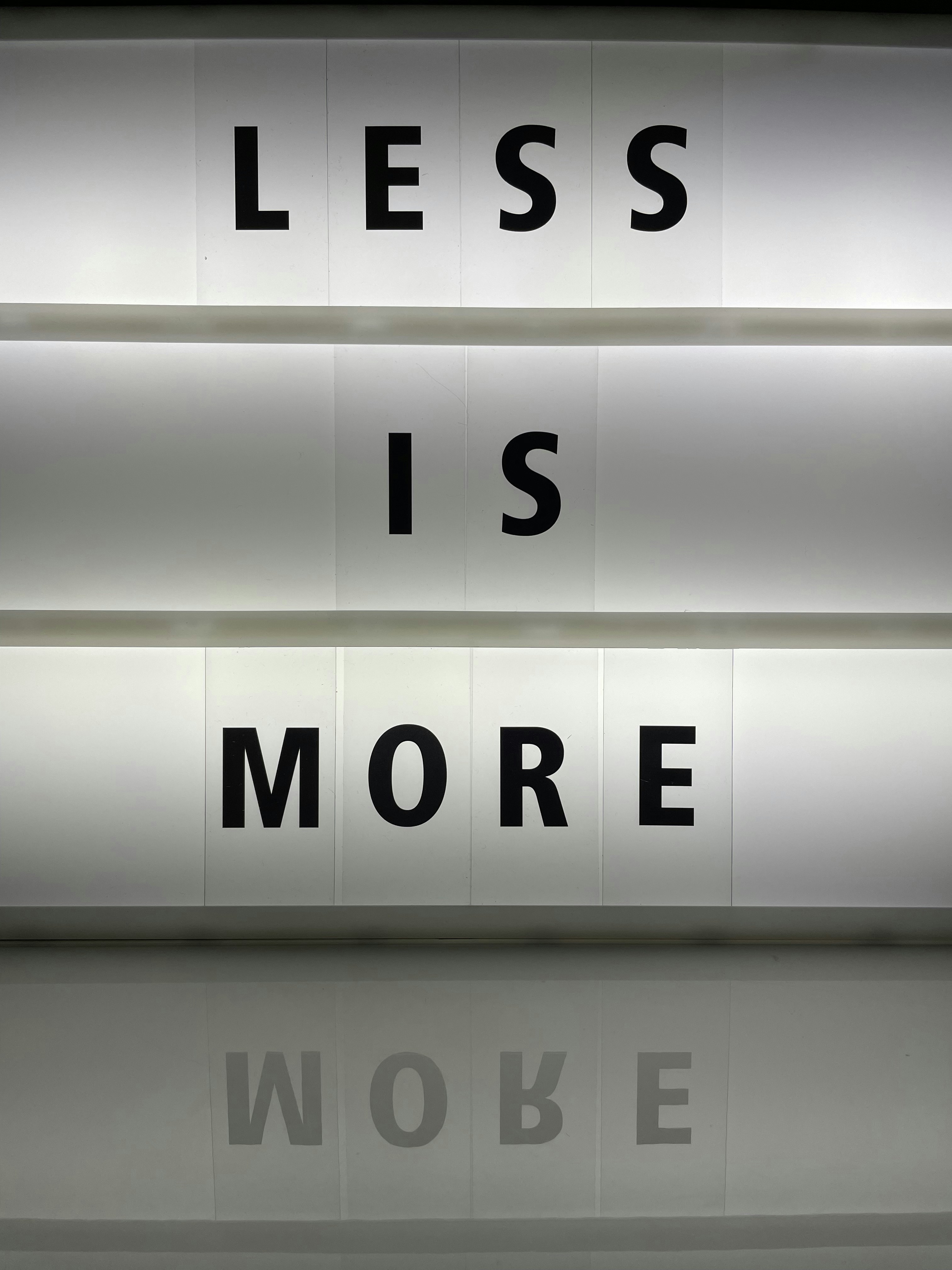 Black letters spelling 'LESS IS MORE' against a softly lit background, reflecting on a glossy surface.