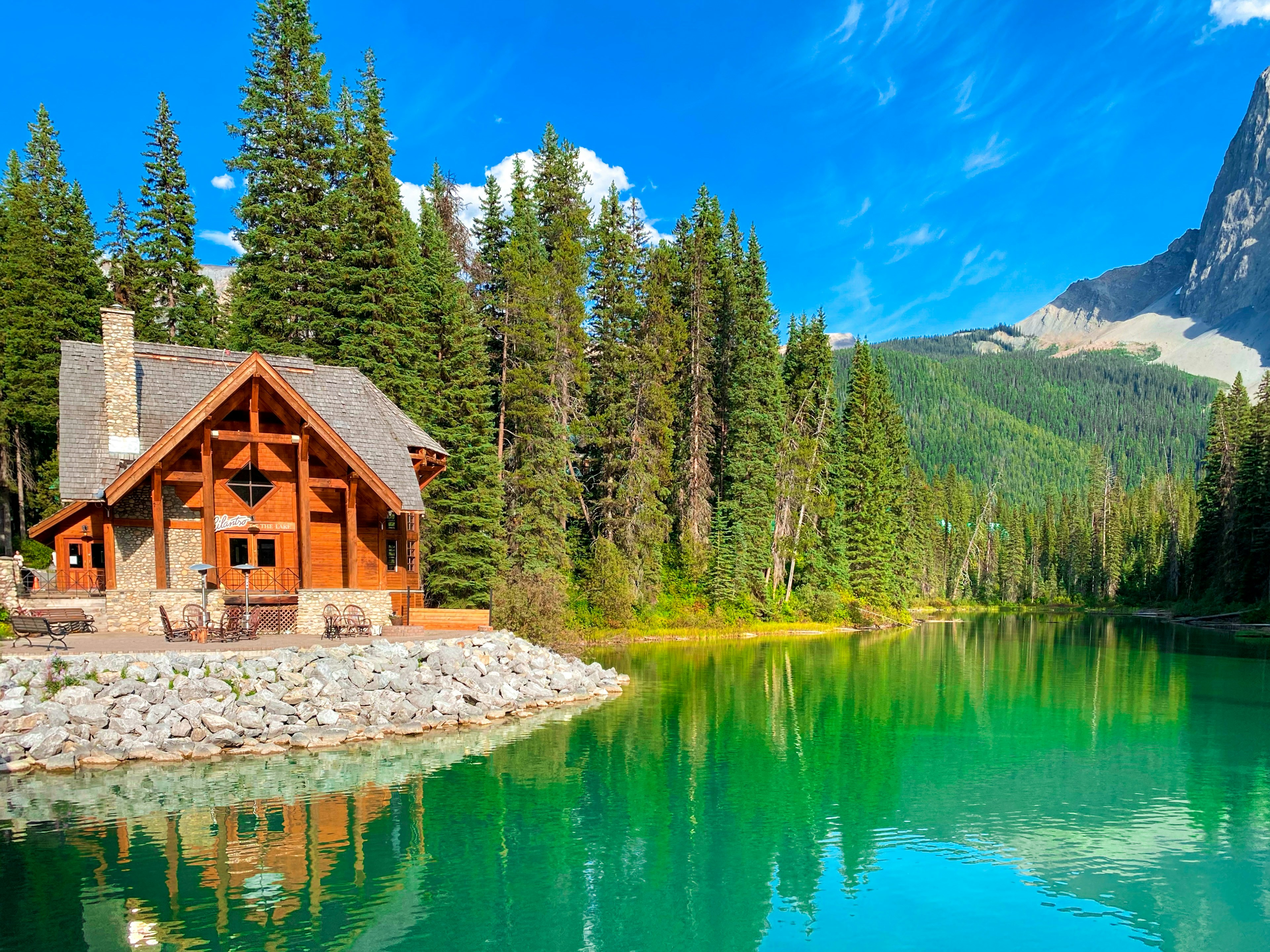 A charming wooden cabin nestled beside a tranquil emerald lake, surrounded by towering pine trees and majestic mountains in the background.