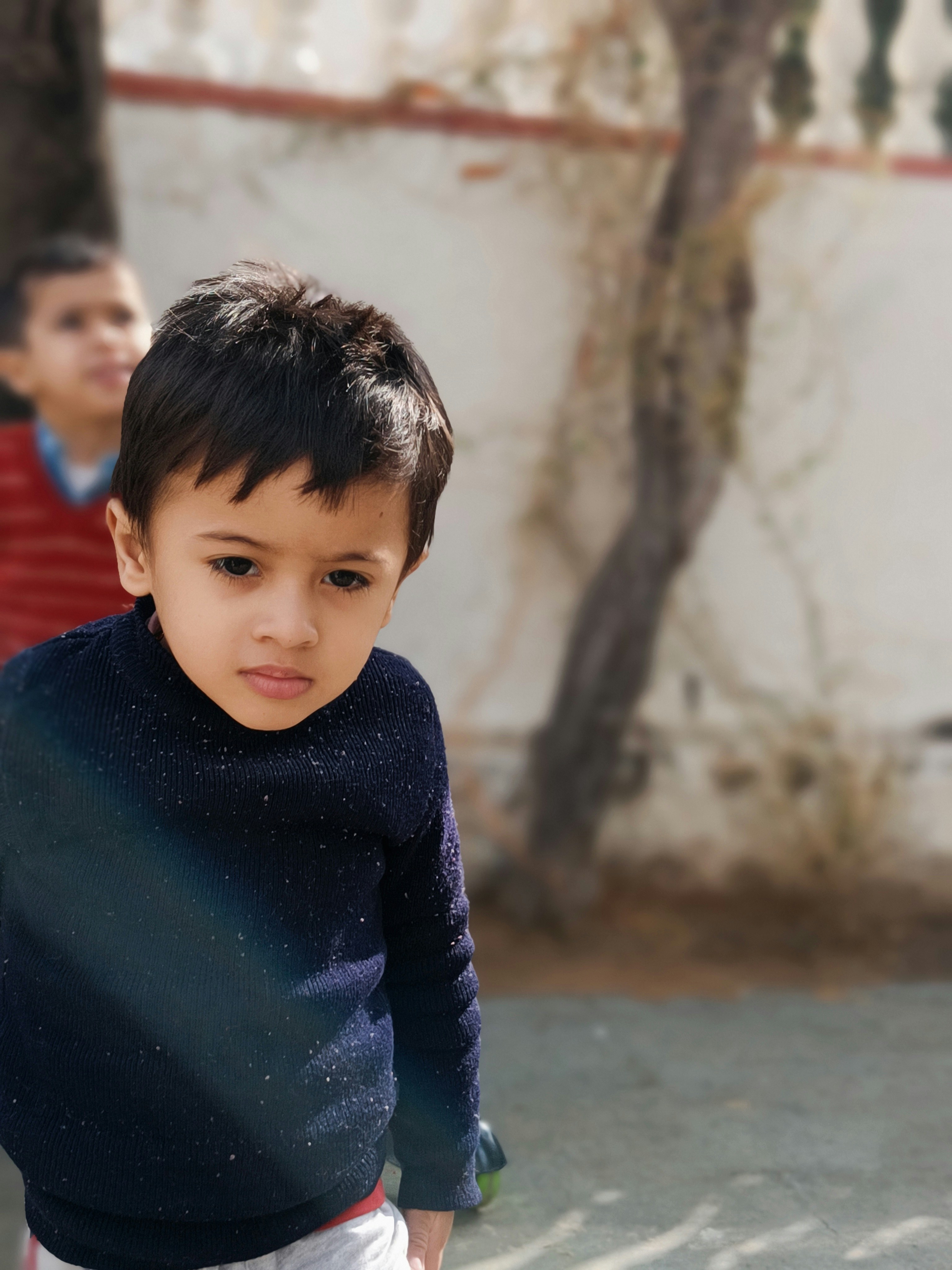Young boy with dark hair and a navy sweater gazes thoughtfully, with a blurred figure in the background. Natural light enhances the scene's warmth.