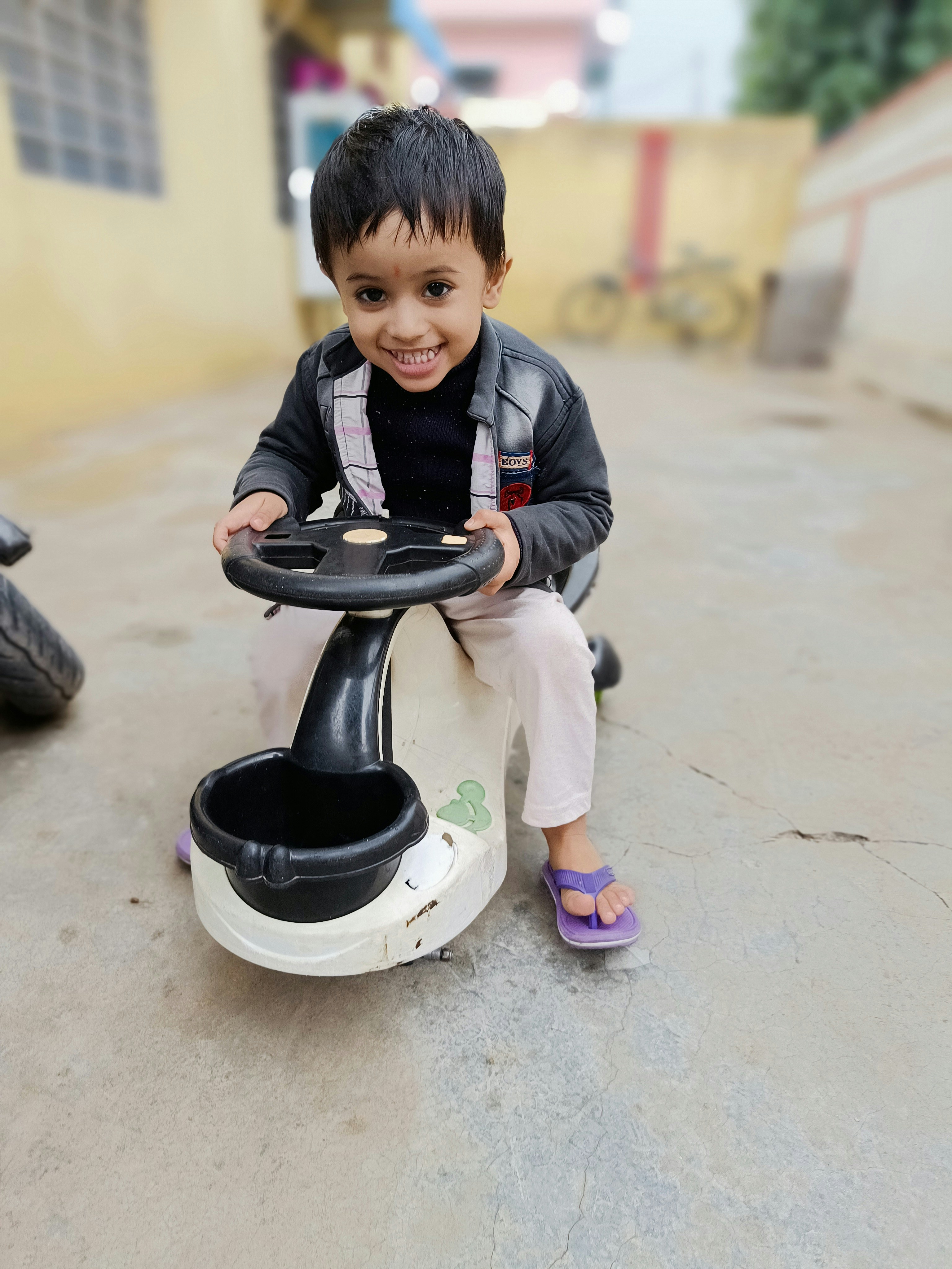 Smiling young boy rides a white plastic trike in a sunlit alley, background blurred.
