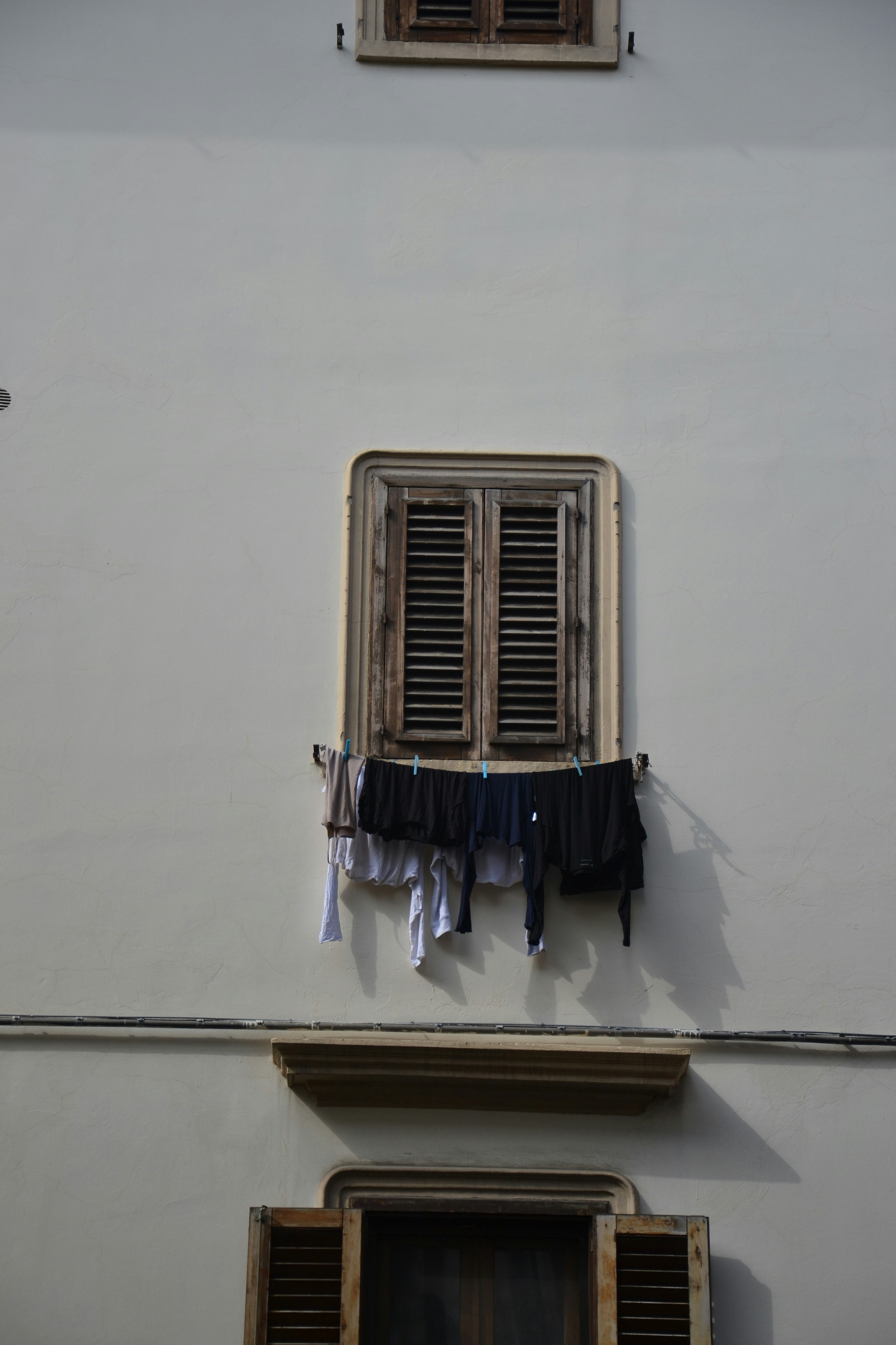 Clothes hang from a clothesline under a wooden shuttered window on a sunlit wall, showcasing everyday life in an urban setting.
