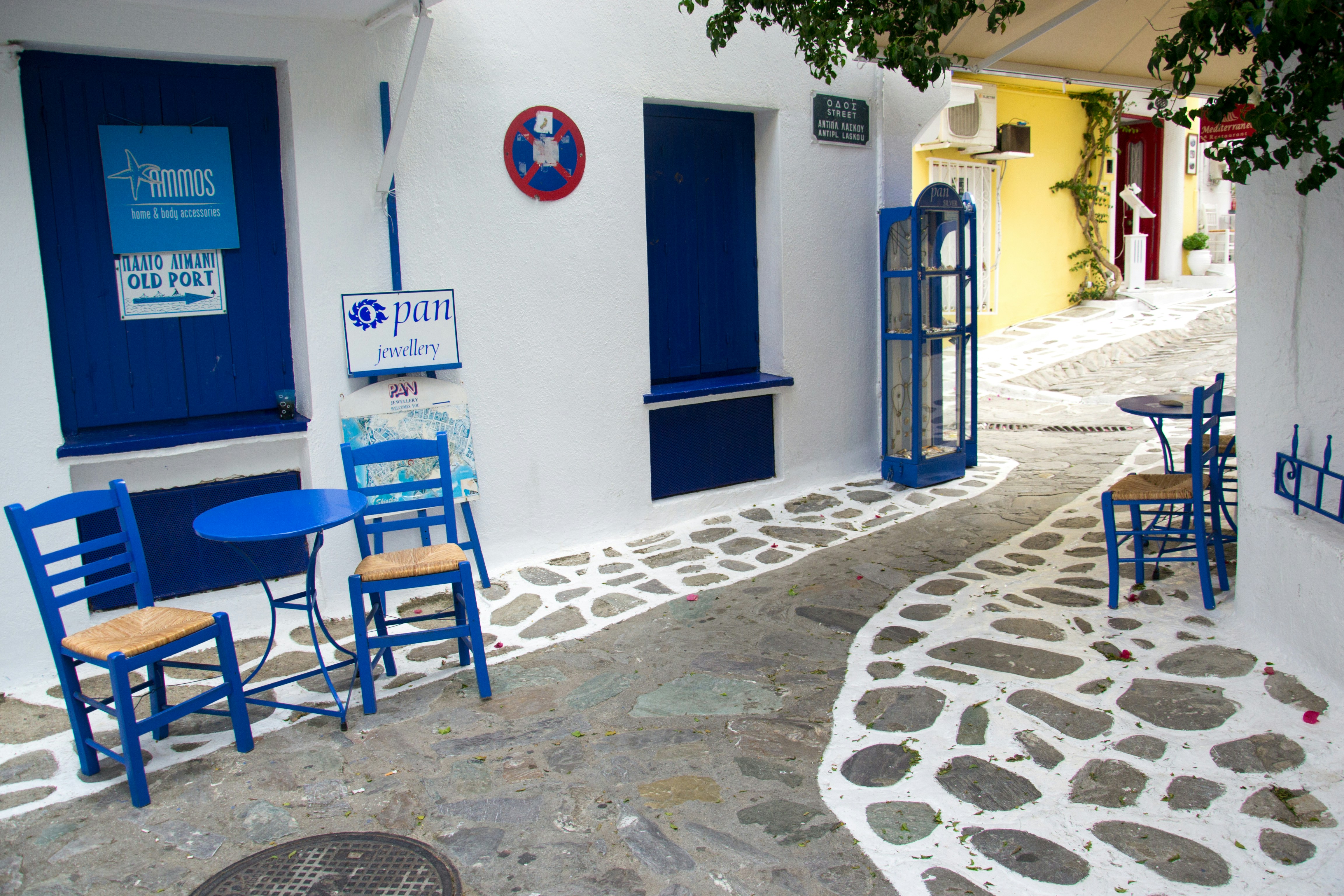 Quaint alley featuring blue furniture and whitewashed walls, leading to vibrant shops in a picturesque coastal setting.