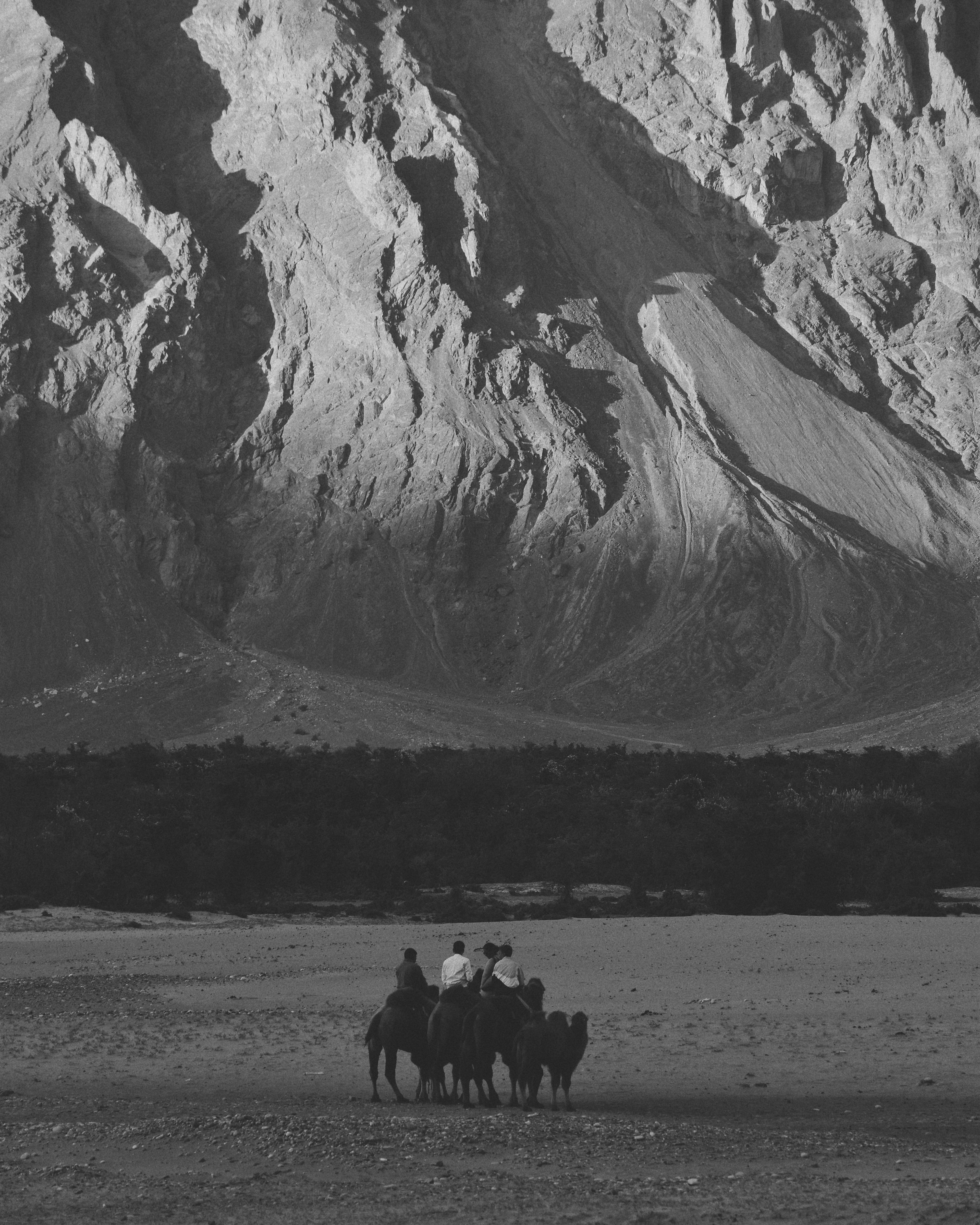 Silhouetted riders on horseback traverse a barren landscape, framed by towering mountains in the background. The monochrome palette enhances the dramatic interplay of light and shadow.